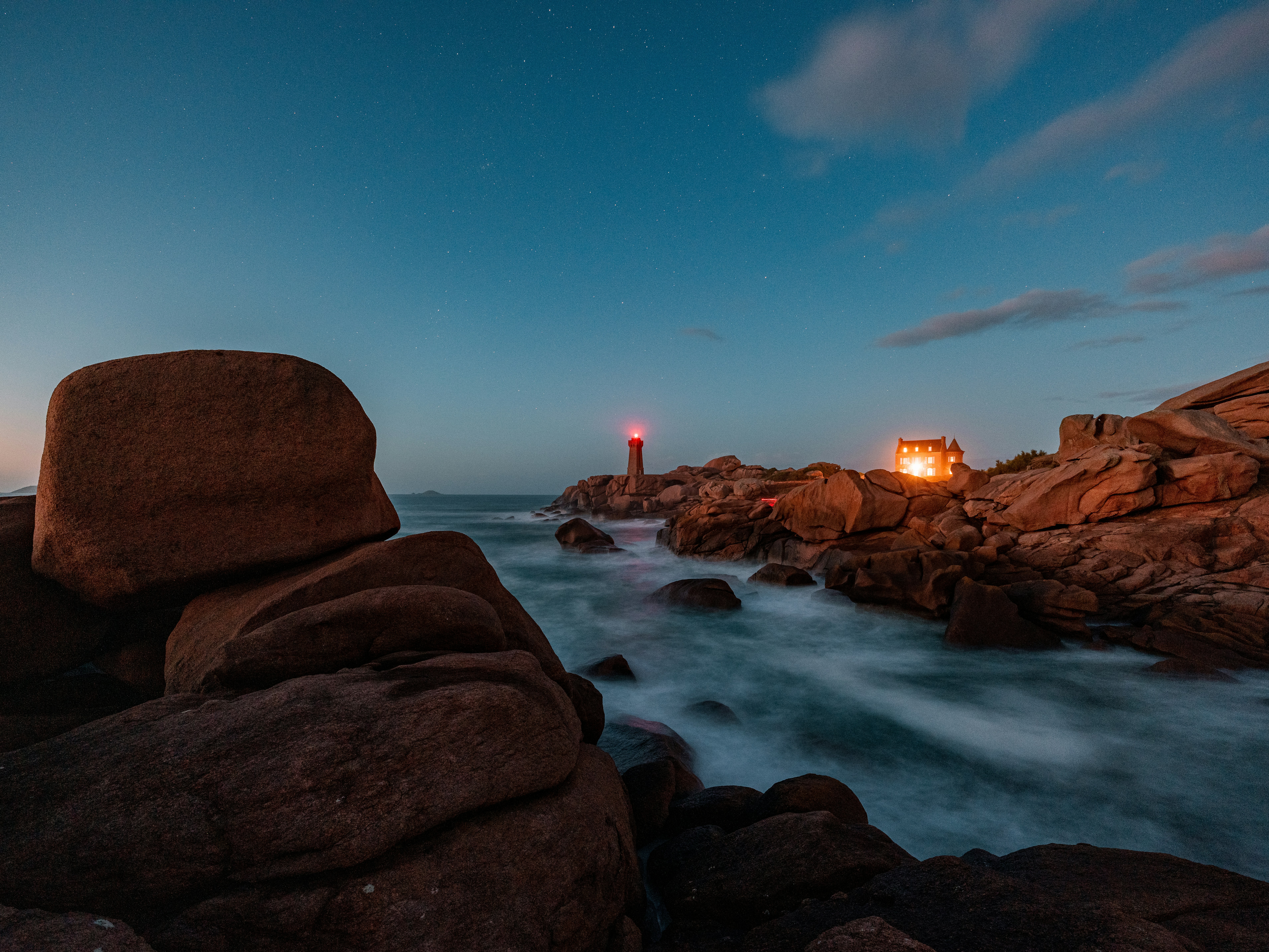 Lighthouse illuminating rocky coastline as waves crash against the shore during twilight.