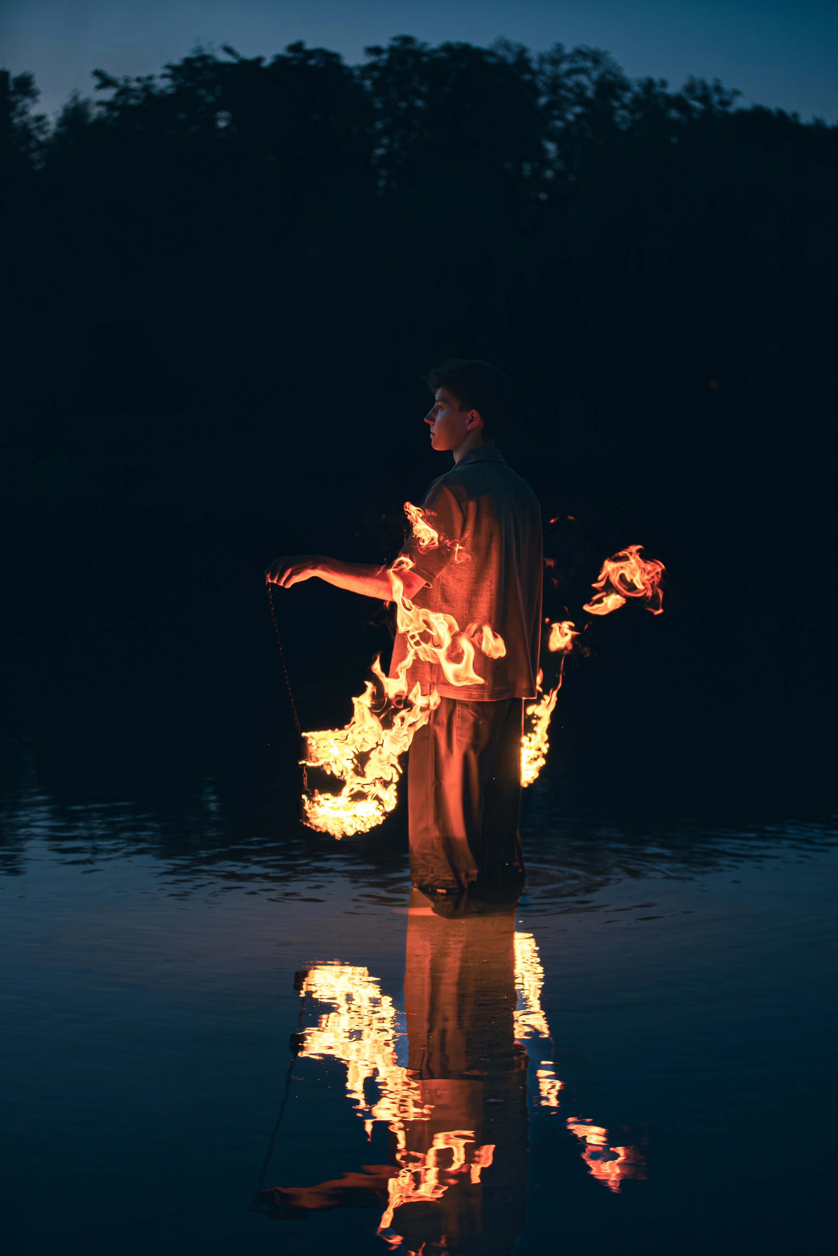 A figure stands in water, holding a flaming object, creating a striking contrast against the darkening sky. The flames reflect on the water's surface, enhancing the dramatic atmosphere.