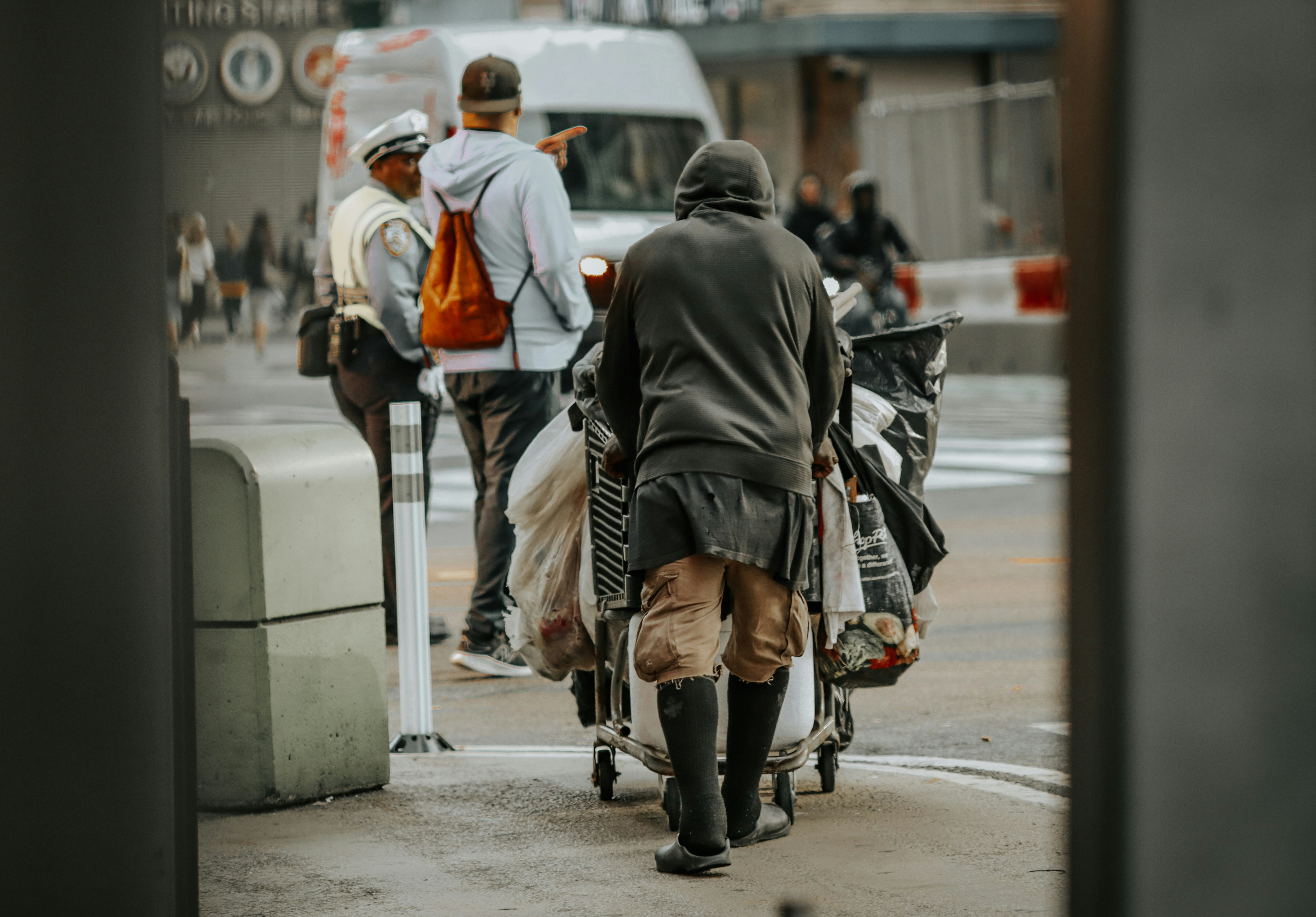 A person pushing a cart filled with belongings navigates a bustling urban street, highlighting the challenges faced by individuals in the city. The scene captures a moment of everyday life amidst the backdrop of city activity.
