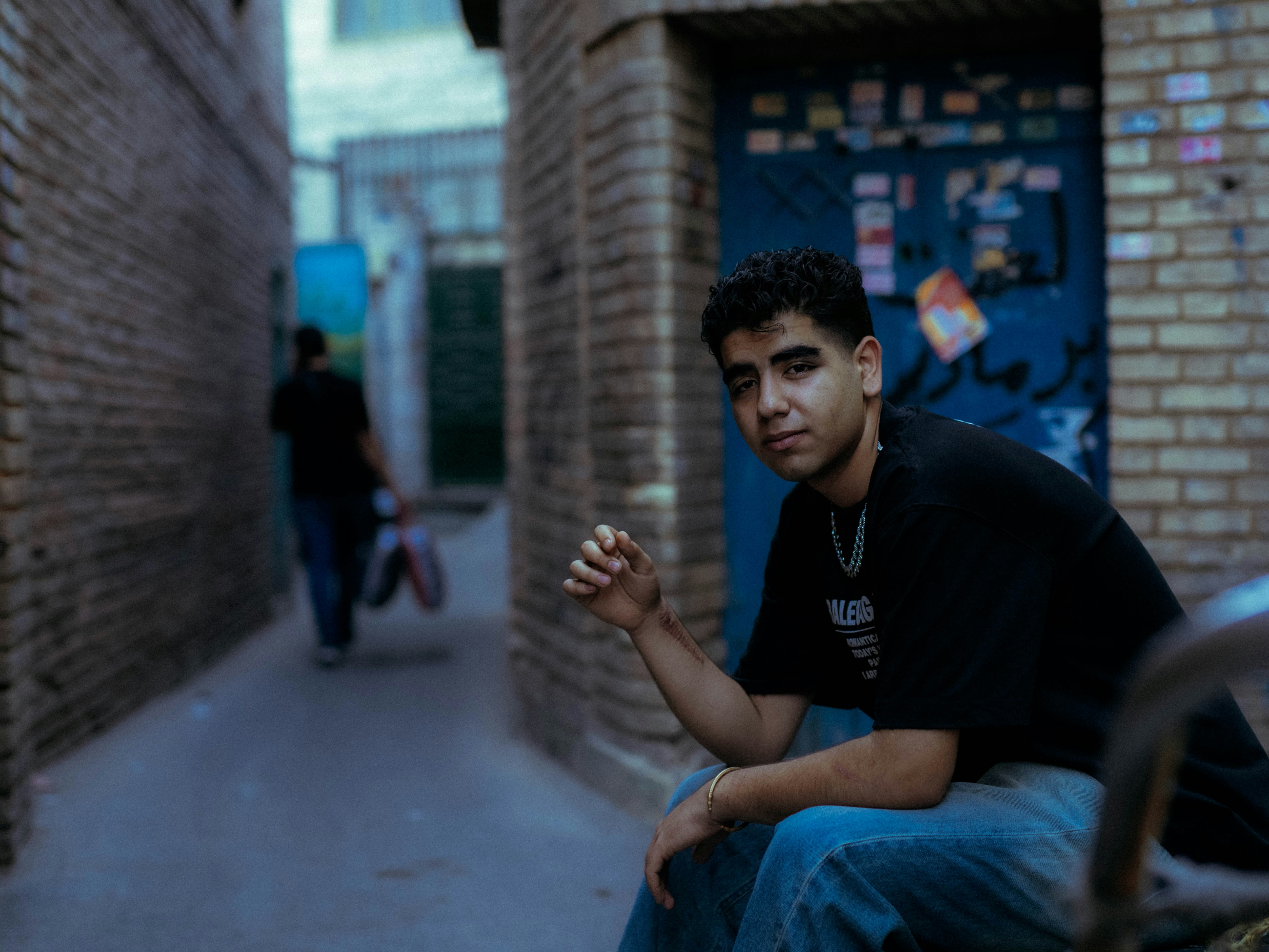 Young man seated casually in an alley, exuding a relaxed vibe amidst the urban backdrop. The scene captures the essence of street life.