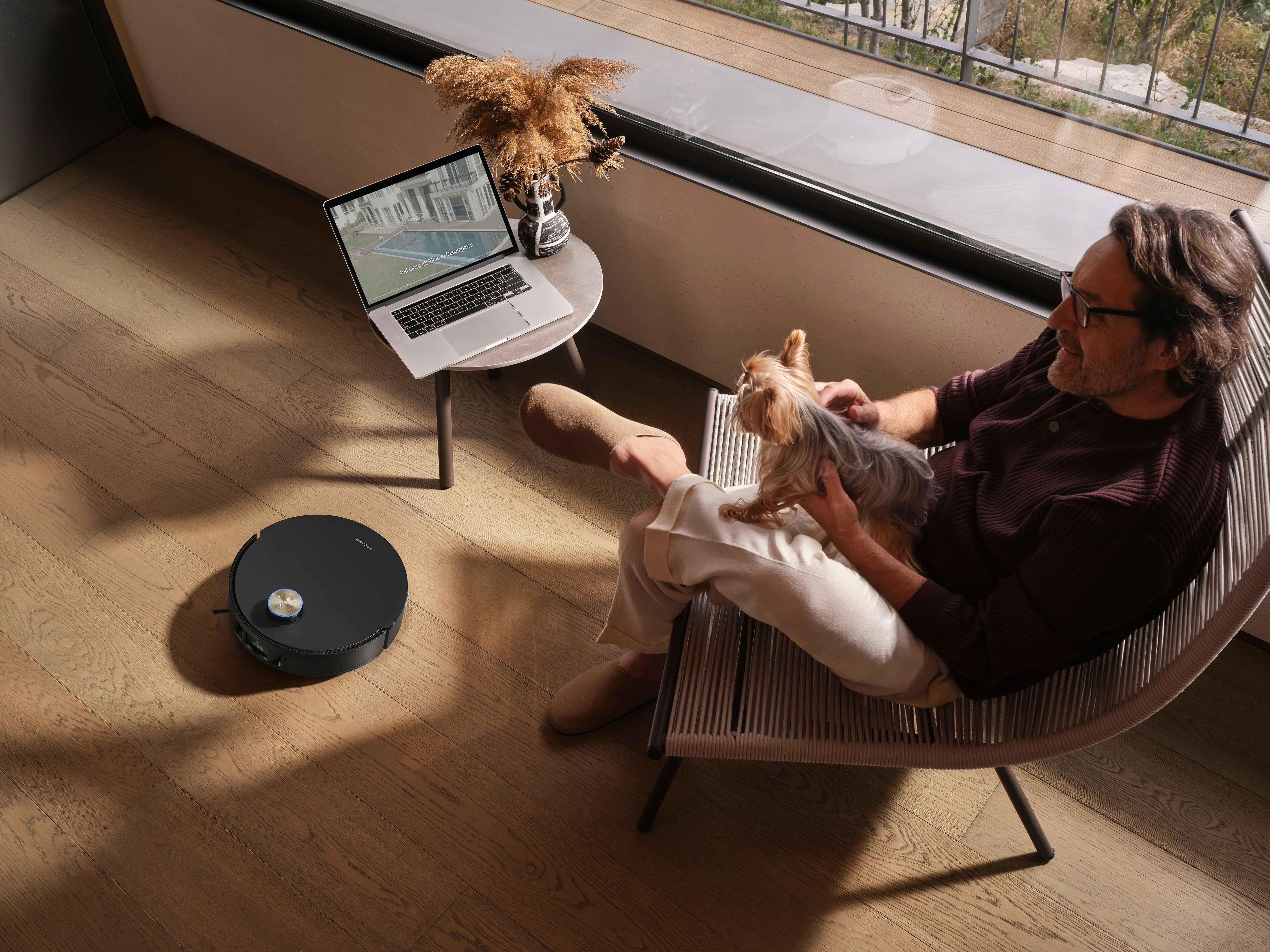 A man relaxes in a stylish chair while holding a small dog, with a laptop open beside him and a robotic vacuum cleaning the floor.