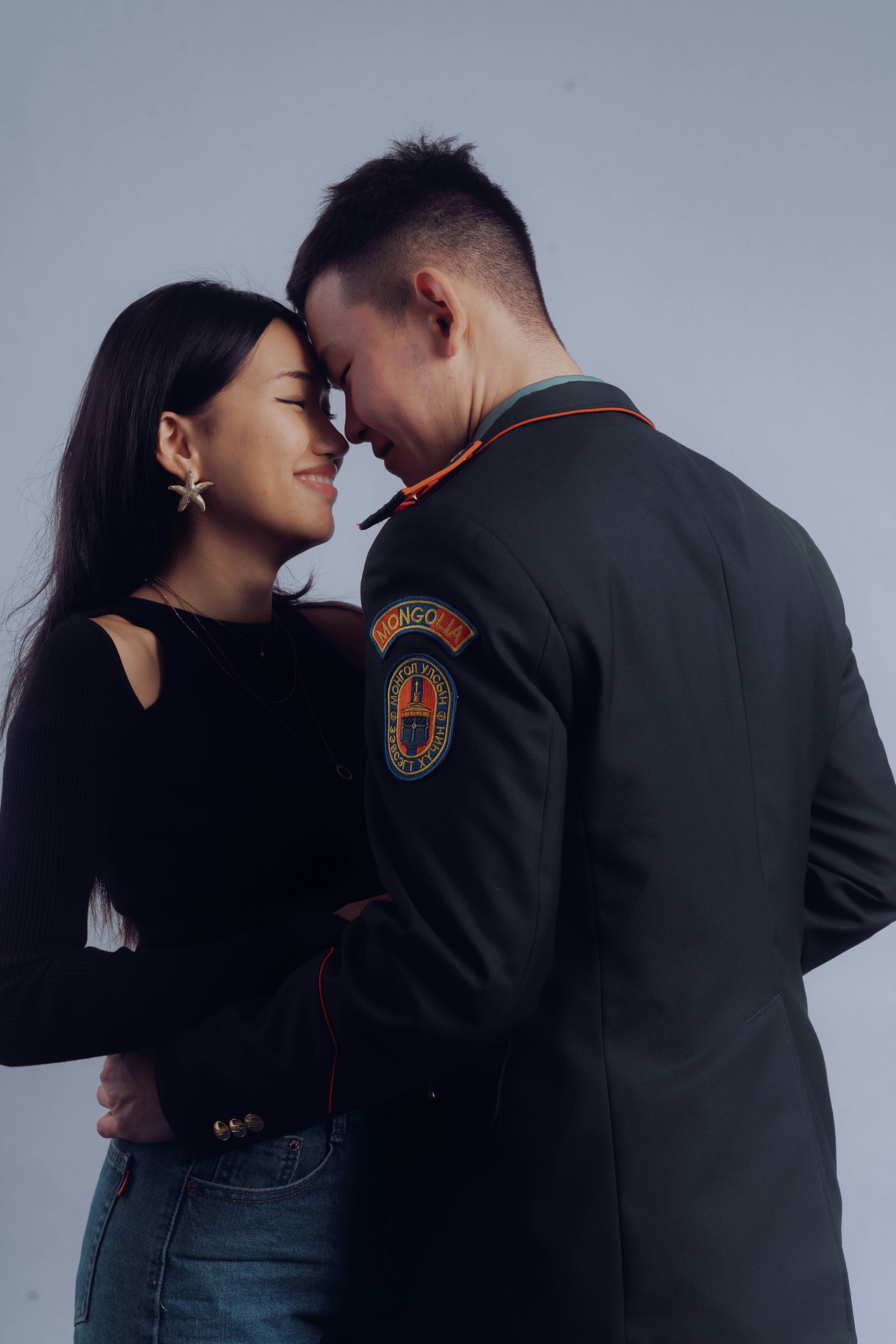 Couple sharing an intimate moment, with the woman in a black top and the man in a military uniform. The soft lighting enhances their affectionate expressions.