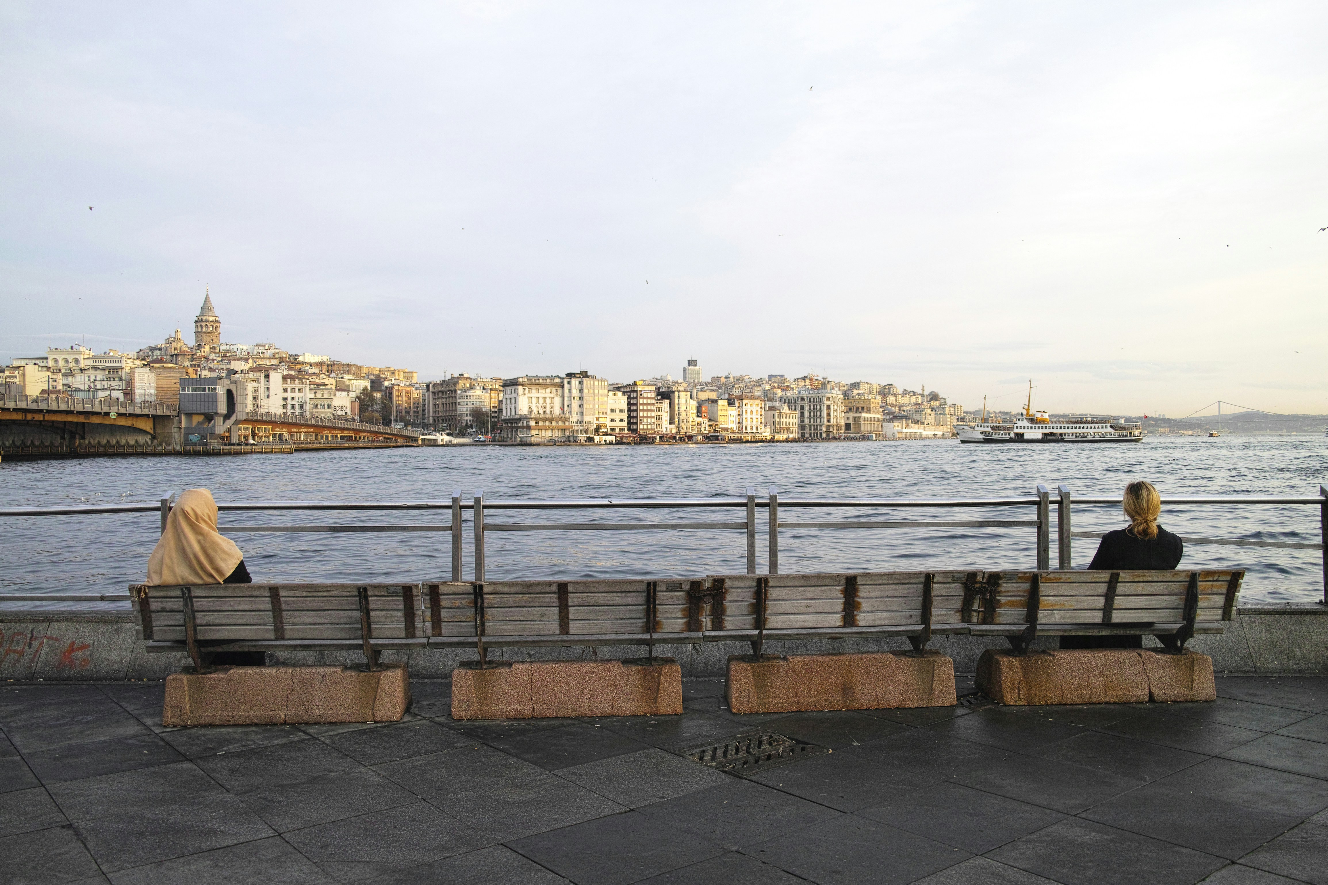 Two women with different cultures sitting on a bench in Istanbul. Contrast of tradition and modernity in city view | Two people sit on a bench overlooking a city skyline.