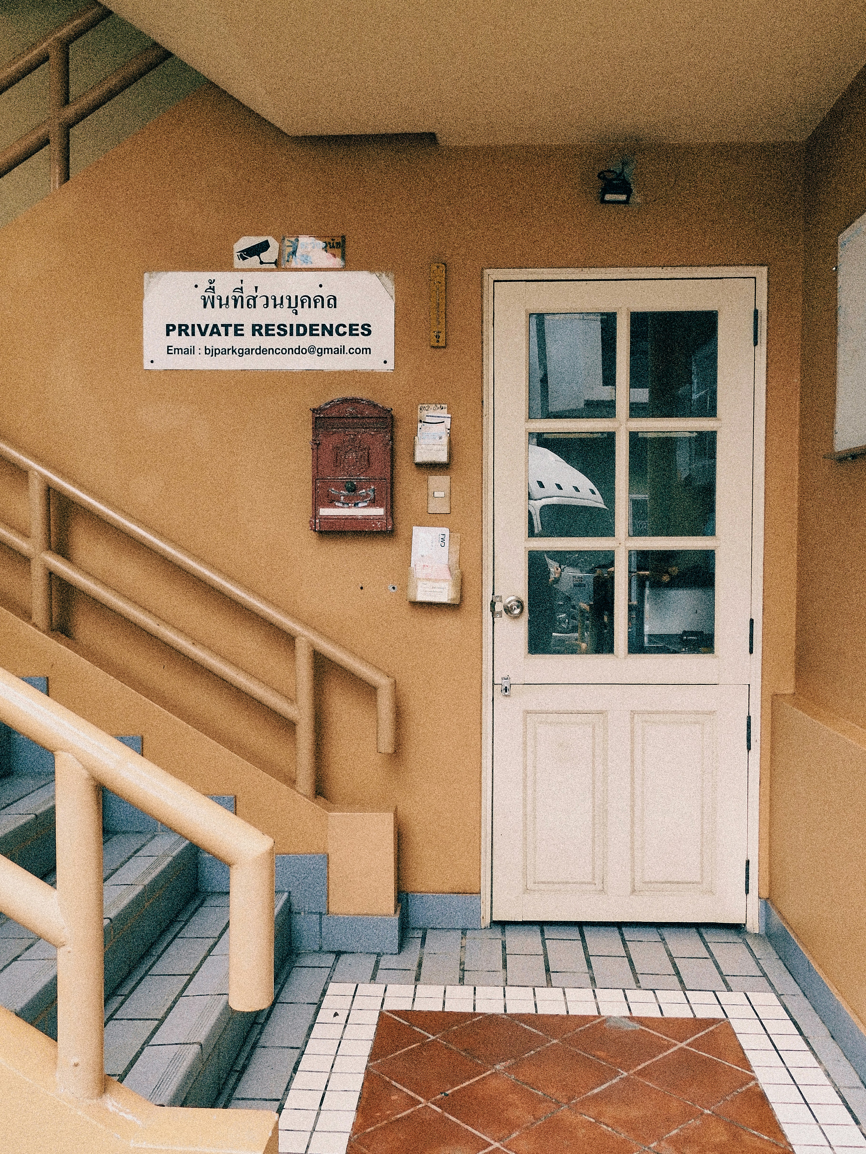 Entrance of a private residence in Thailand, with a vintage red mailbox, cream-colored door, and tiled floor. The warm tones of the building contrast with the clean white details, giving the photo a nostalgic, cinematic feel. The sign above the door reads “Private Residences” in both Thai and English, adding a sense of cultural and urban authenticity. | Entrance to private residences with stairs and door