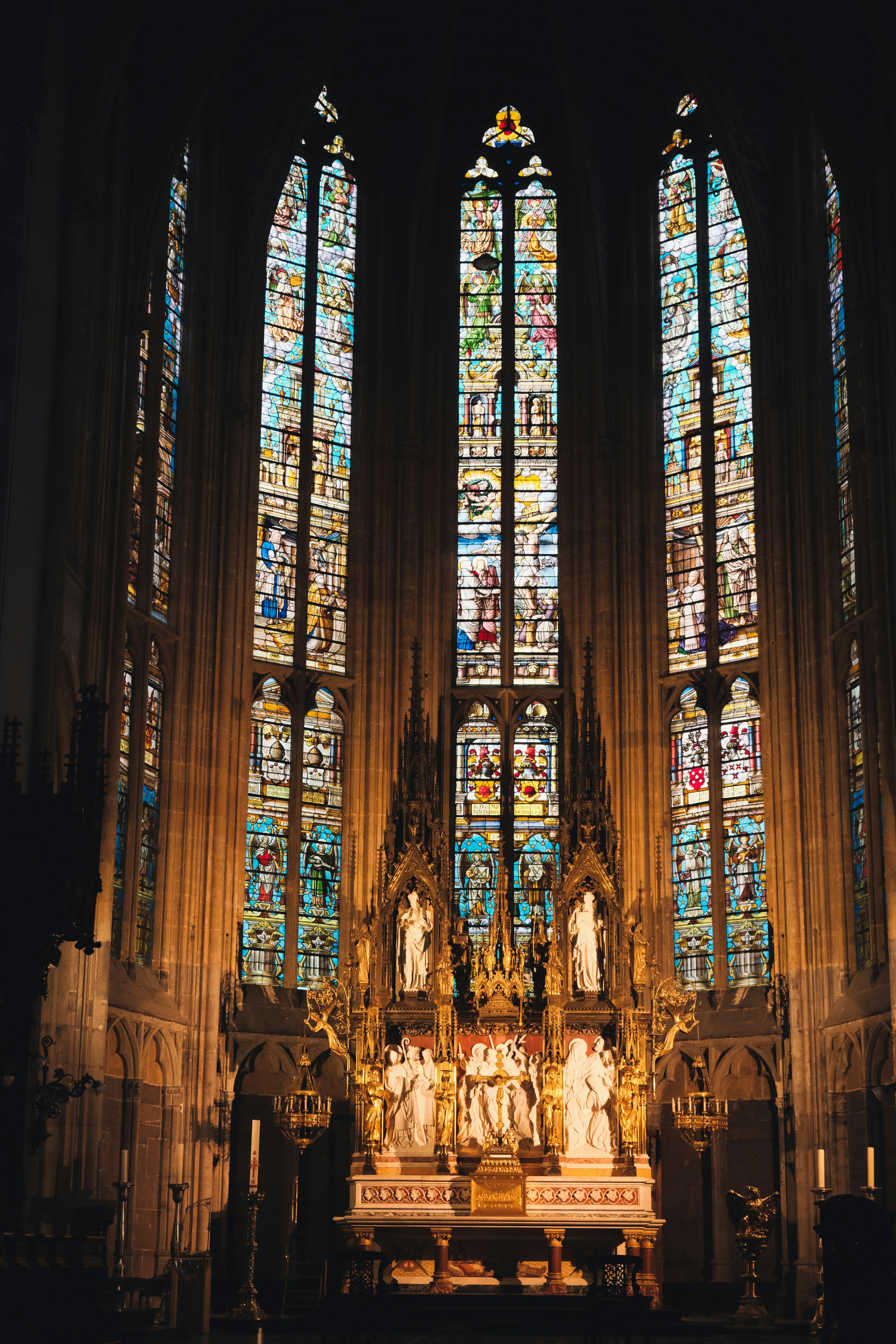 Intricate stained glass windows illuminate a majestic altar, showcasing fine sculptures and ornate details in a sacred space.