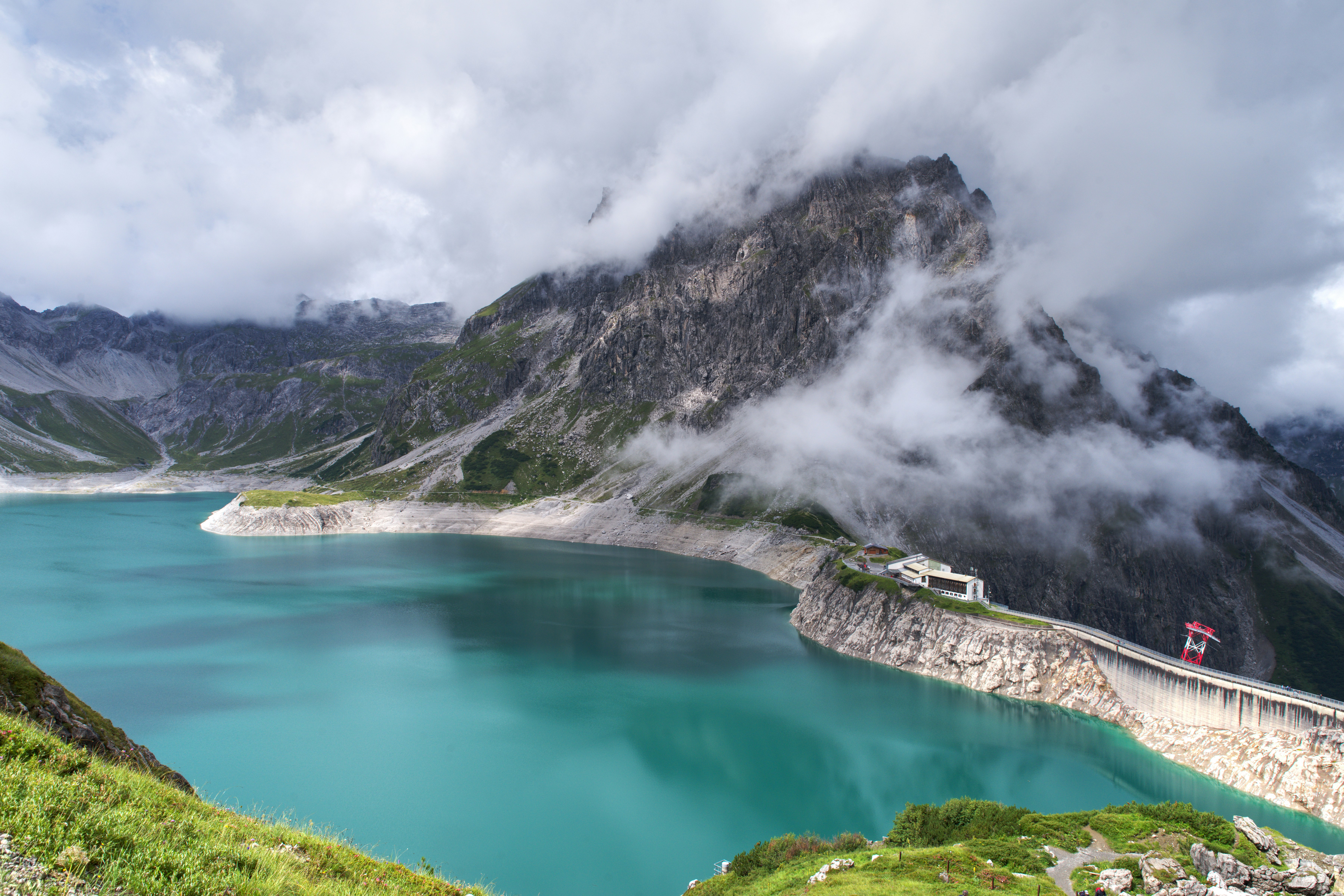 Turquoise lake reflecting dramatic mountain peaks under a cloudy sky, with a dam and structures along the shore.