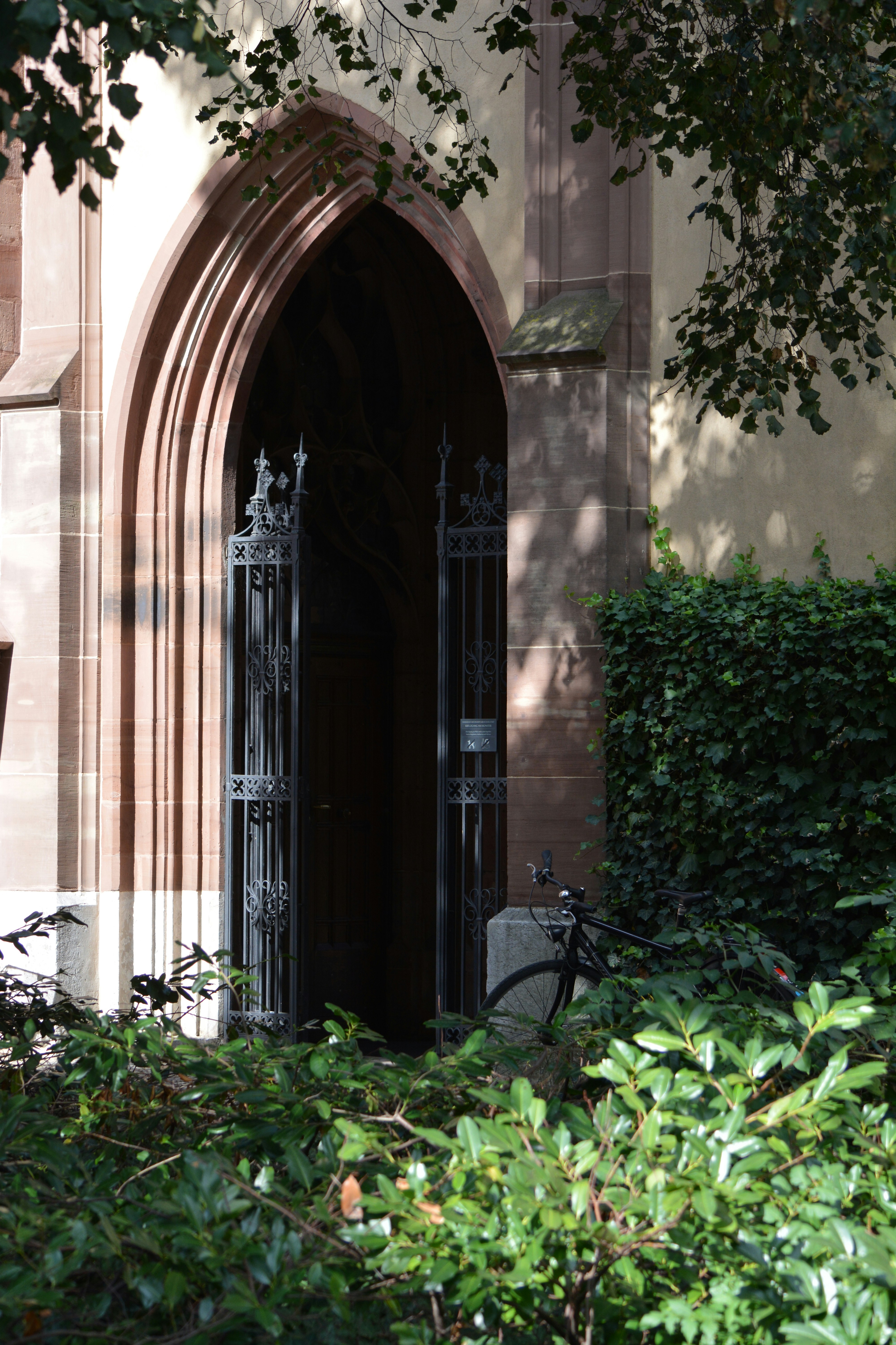 Intricate wrought iron gates stand before a shadowed doorway, framed by lush greenery and ancient stone architecture.