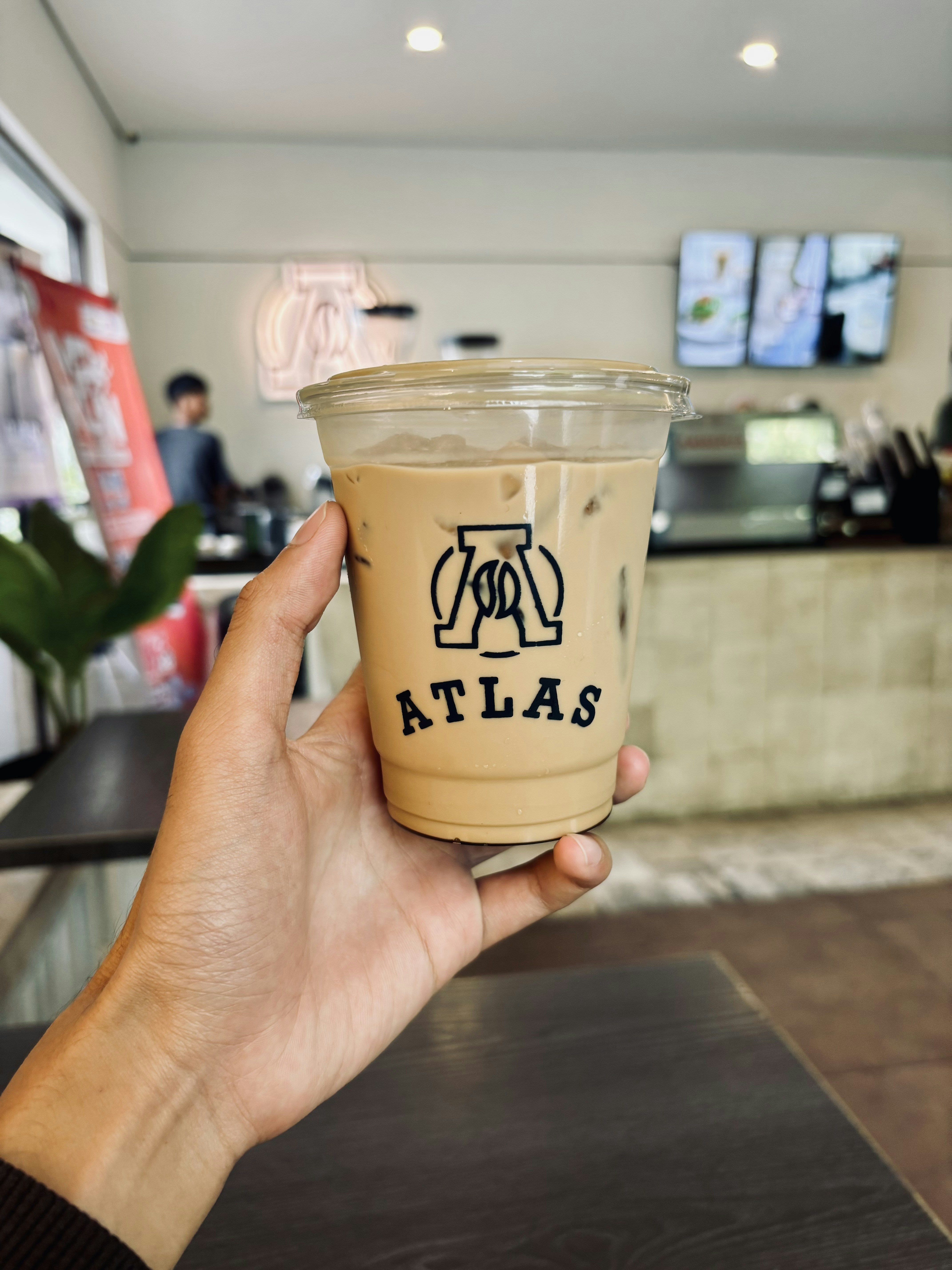 Hand holding a cup of iced coffee from Atlas, featuring a prominent logo, set against a cozy café backdrop.