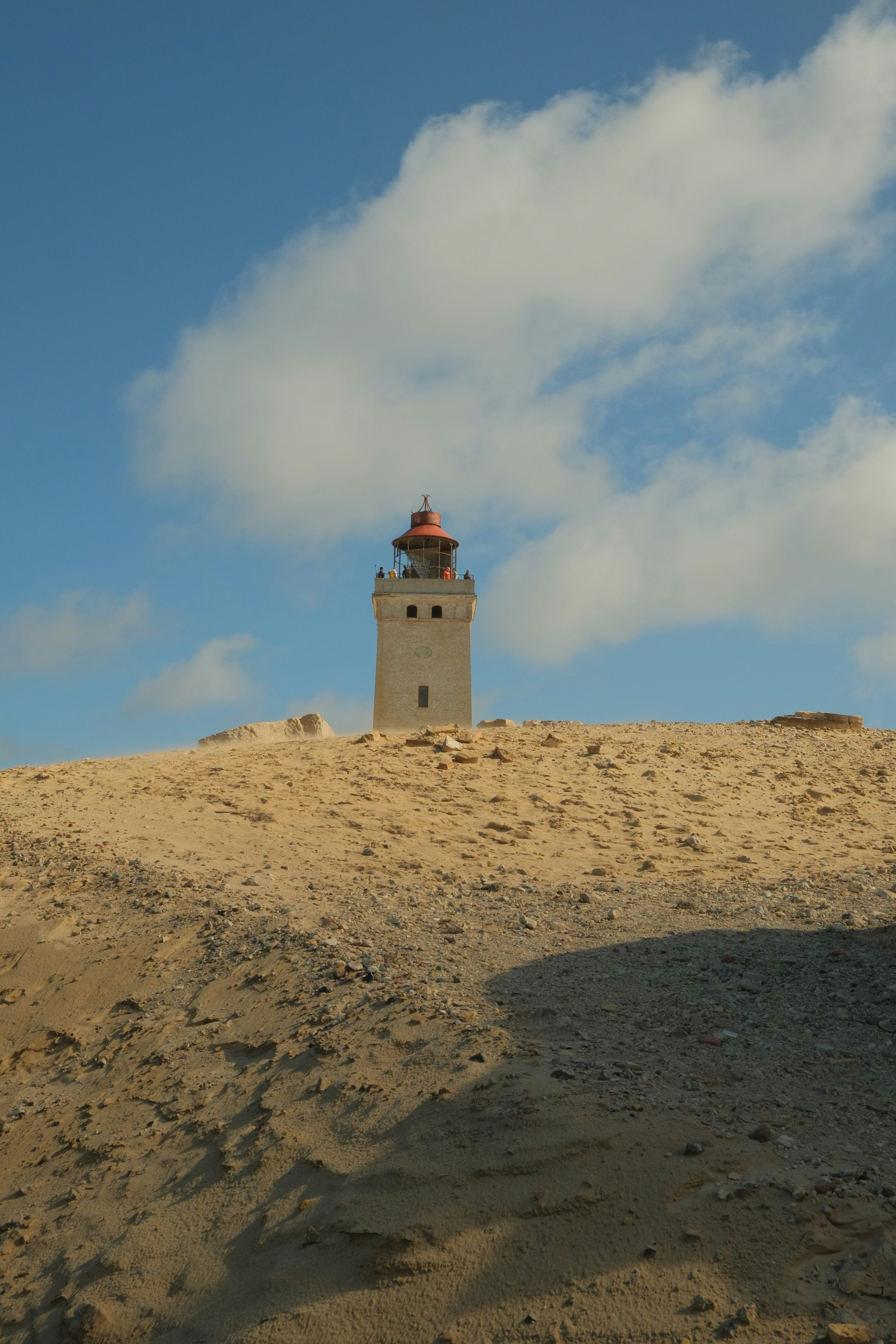 A lighthouse stands on a sandy hill under blue skies.