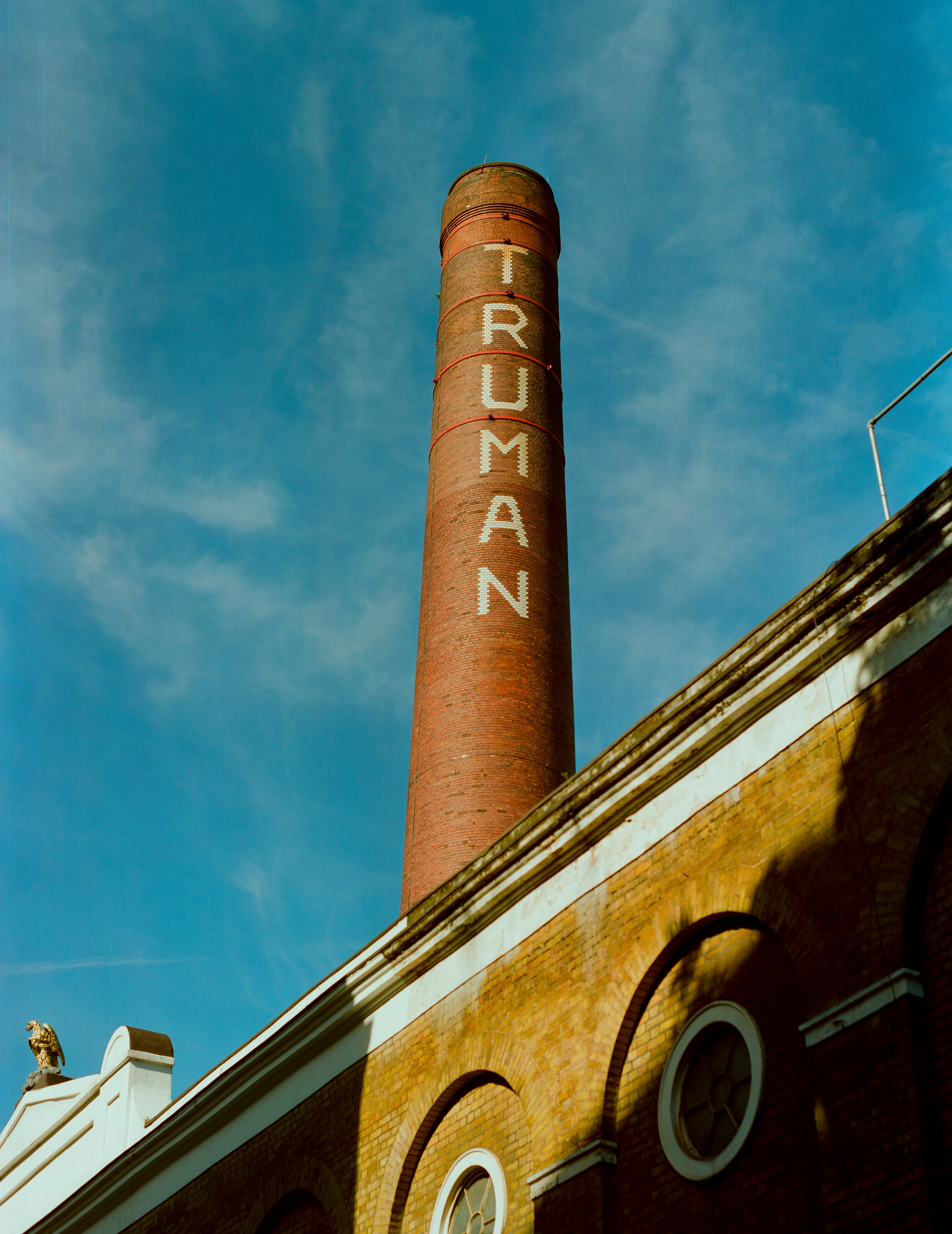 Capturing the iconic Truman Brewery on a vibrant afternoon, this shot exudes nostalgia. | Brick chimney with "truman" text against blue sky.