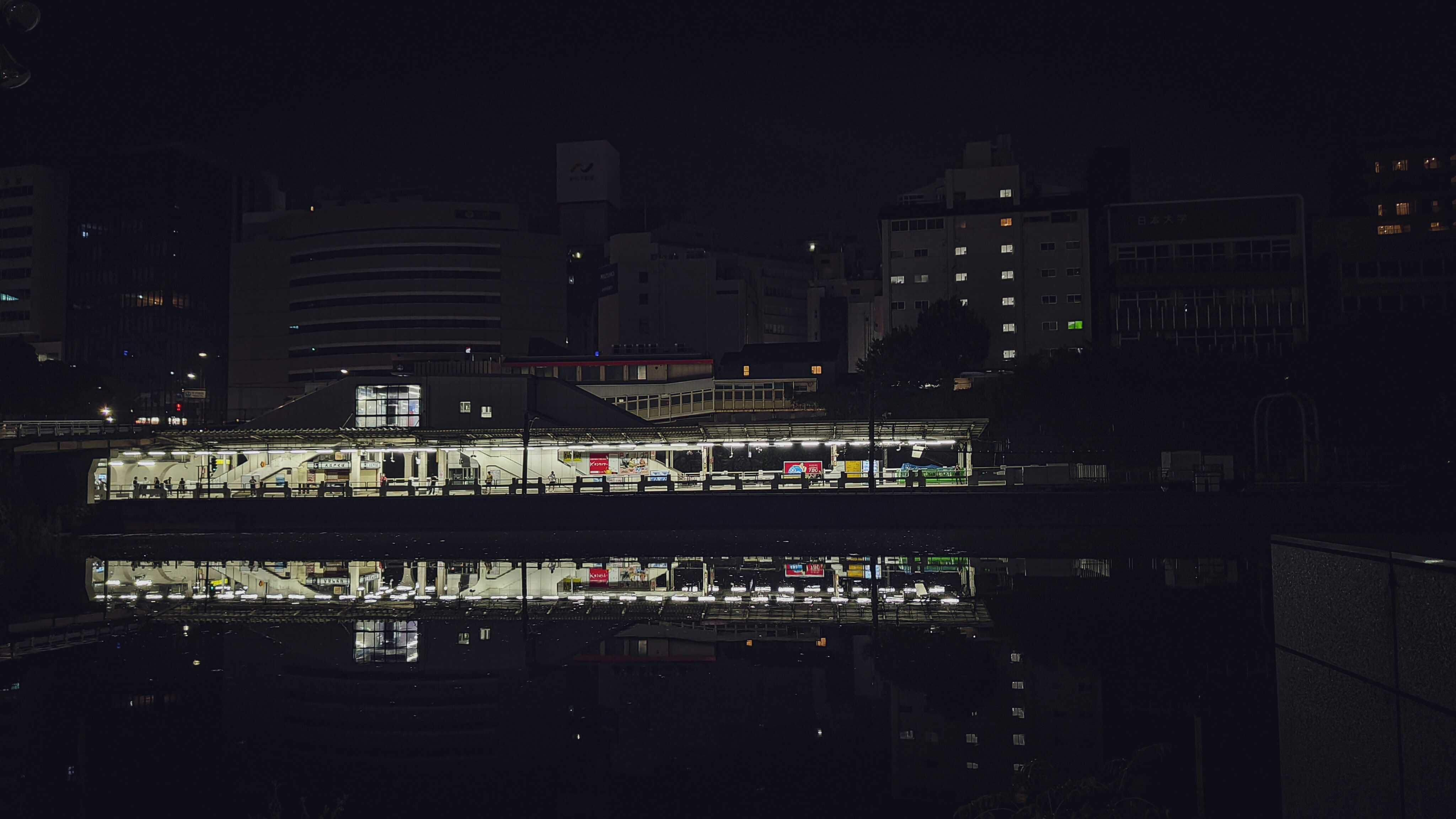 Train station illuminated at night, reflecting on a still body of water, surrounded by urban architecture.