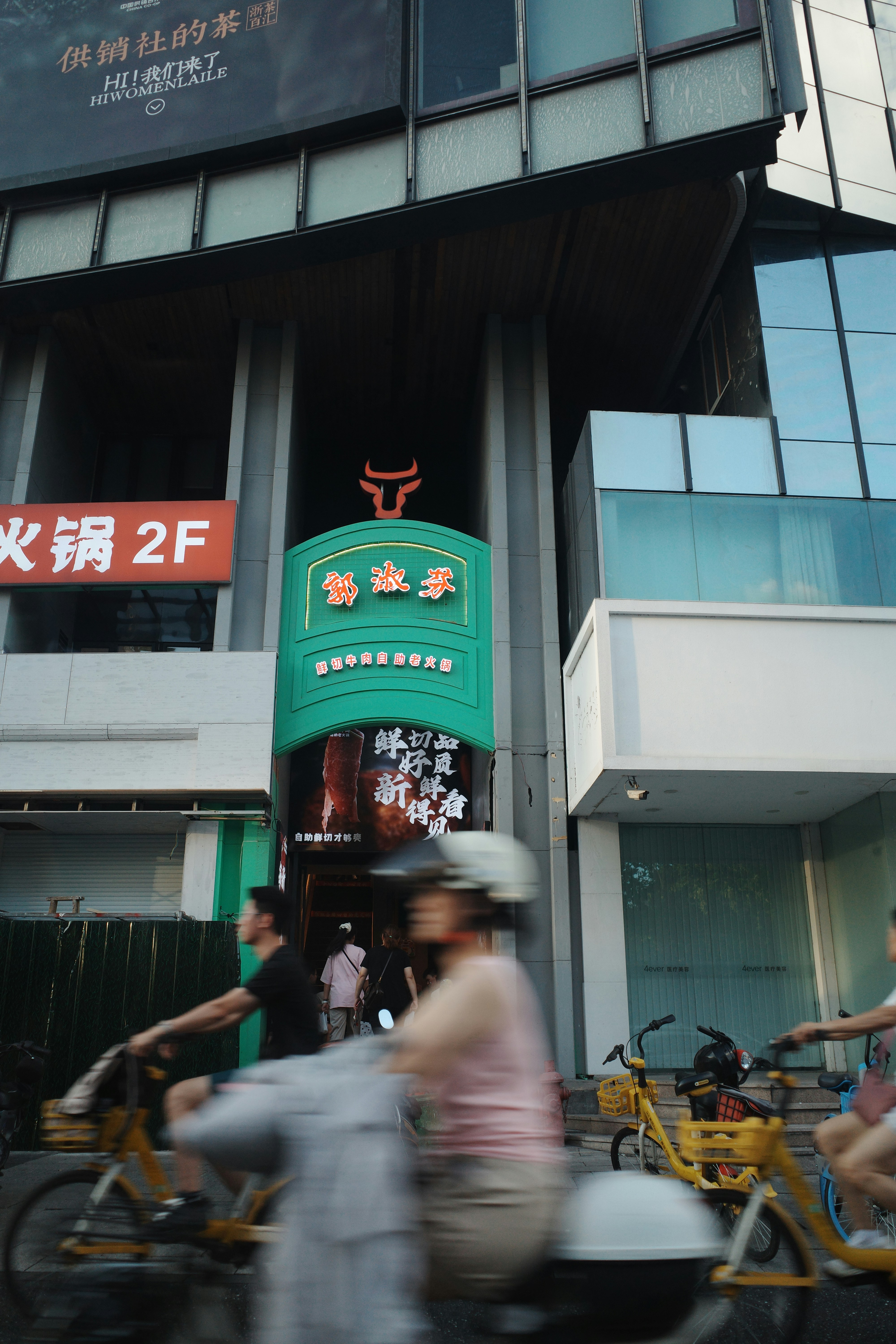 People cycling past a restaurant entrance in a city.