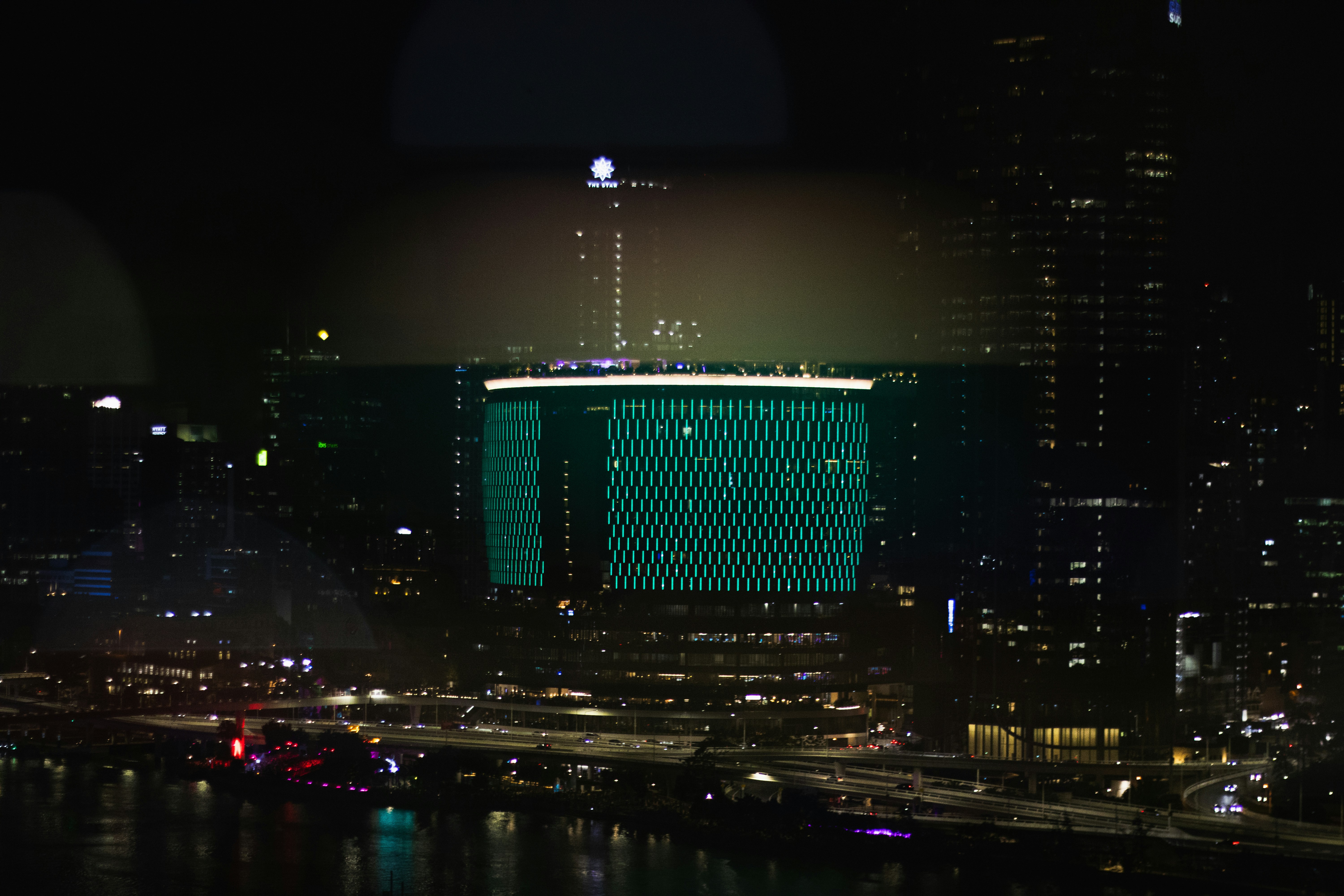 A vibrant building illuminated in turquoise lights, surrounded by a bustling city skyline at night. The reflections on the water enhance the lively atmosphere.