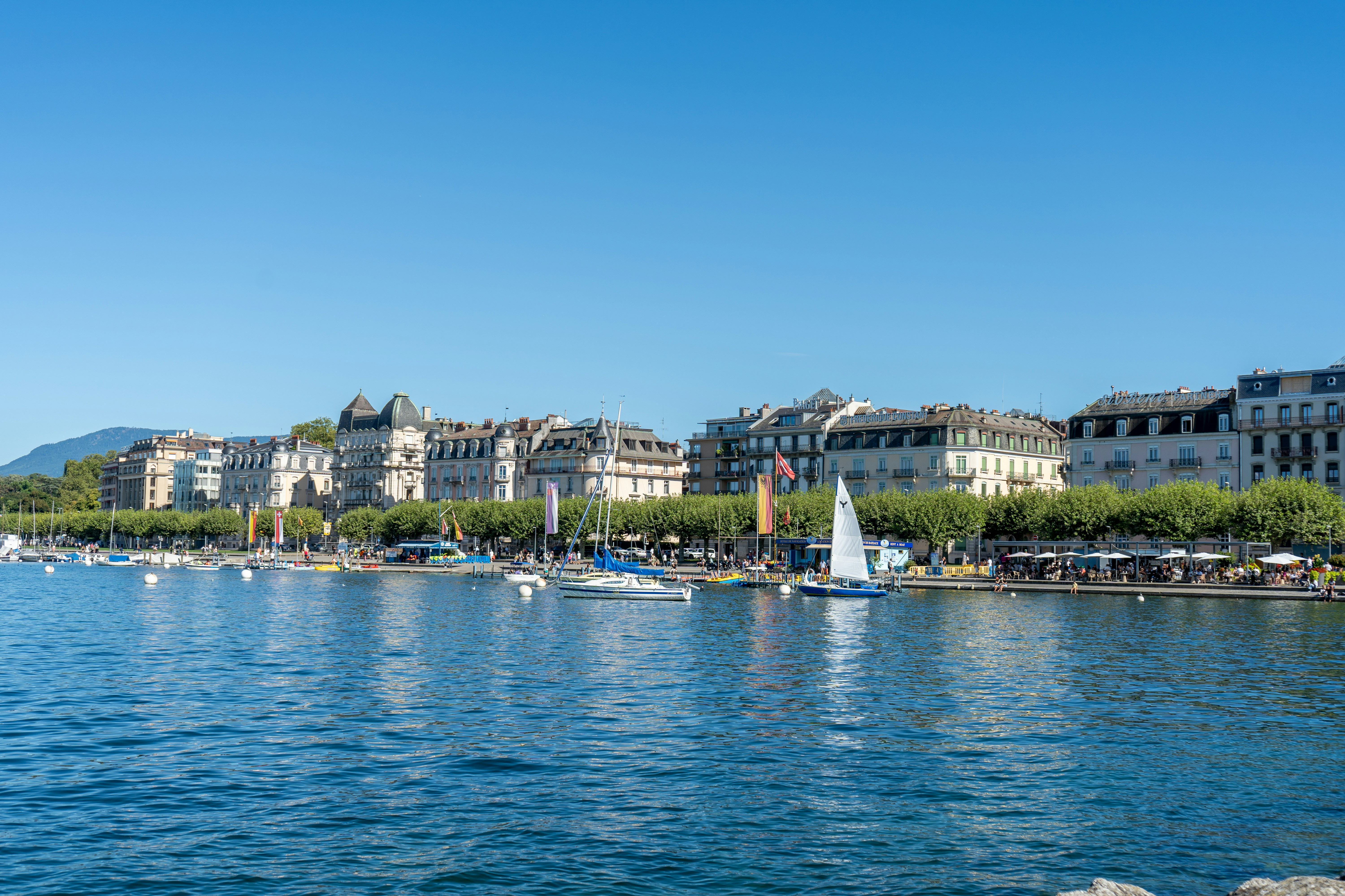 Lively lakeside scene in Geneva featuring charming buildings, sailboats, and vibrant trees along the waterfront.