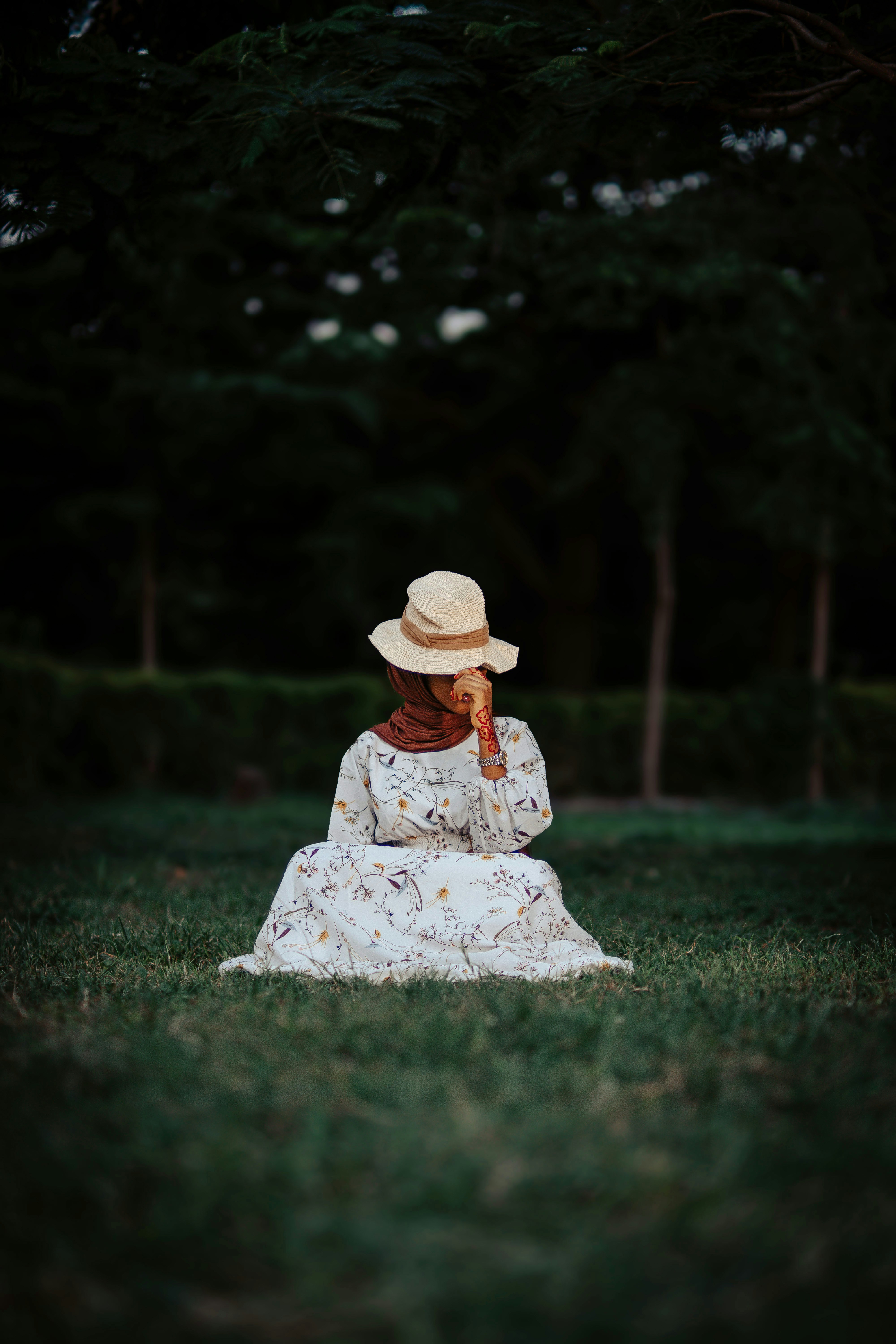 A figure in a floral dress and hat sits peacefully on the grass, holding a flute, surrounded by lush greenery. The composition evokes tranquility and reflection.