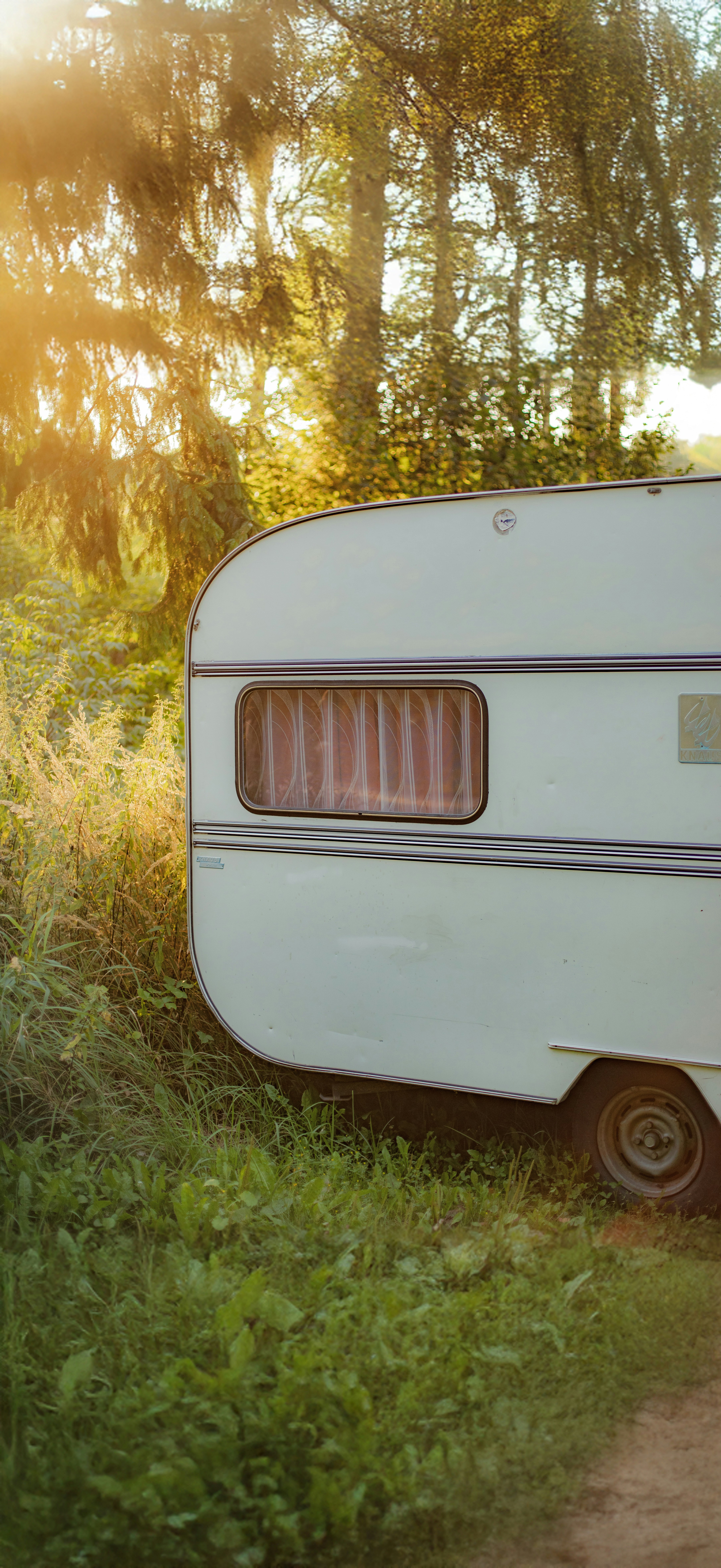 This image captures a vintage caravan resting peacefully in a lush, green woodland clearing. The soft afternoon light filters through the trees, casting a warm glow on the weathered exterior of the camper. The scene evokes a sense of quiet adventure and nostalgic tranquility, perfect for themes of travel, escape, and the simple beauty of nature. | Caravan parked in a grassy, sunlit forest clearing.