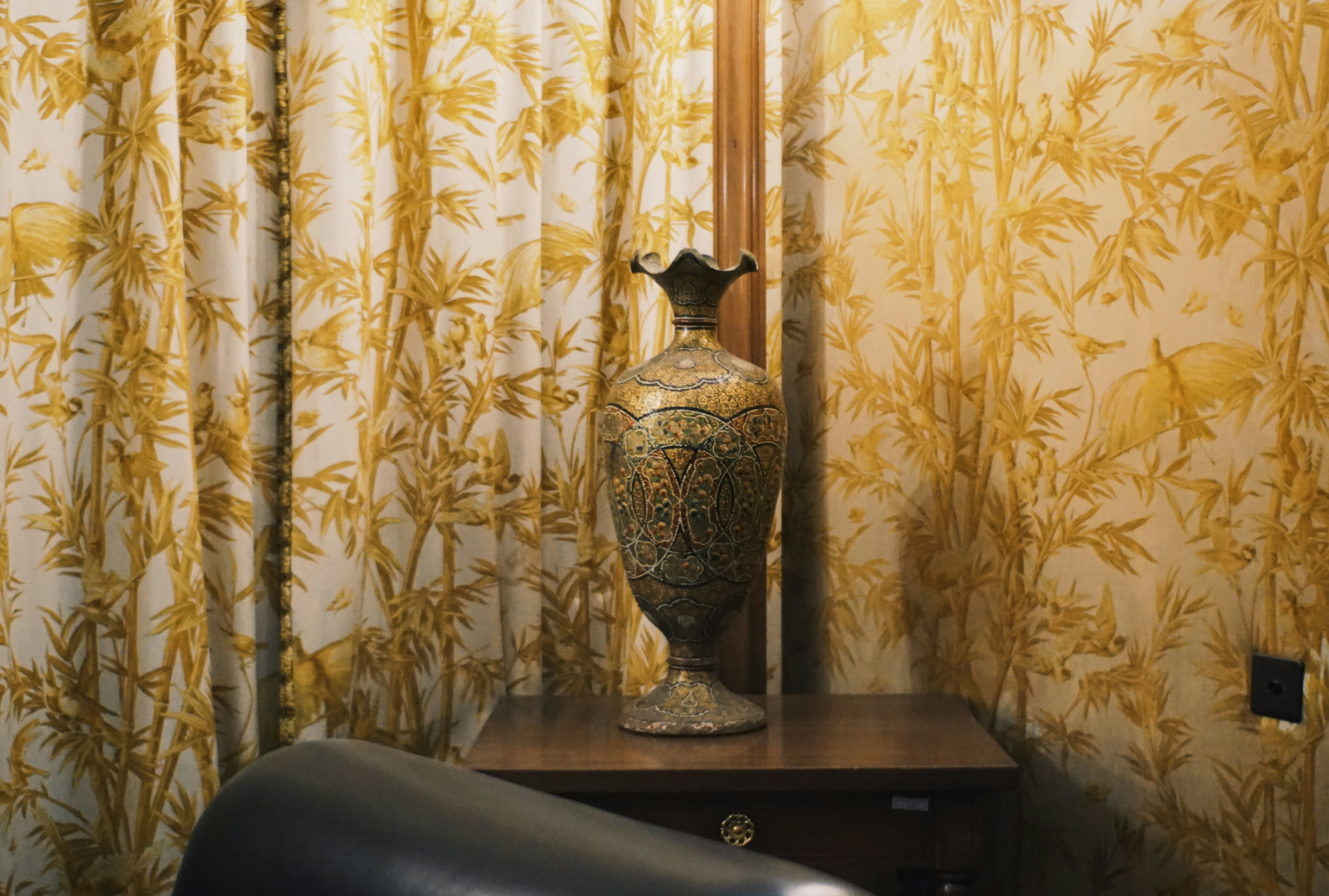 Intricately designed vase resting on a wooden table, surrounded by rich, floral-patterned curtains that evoke a sense of nostalgia.