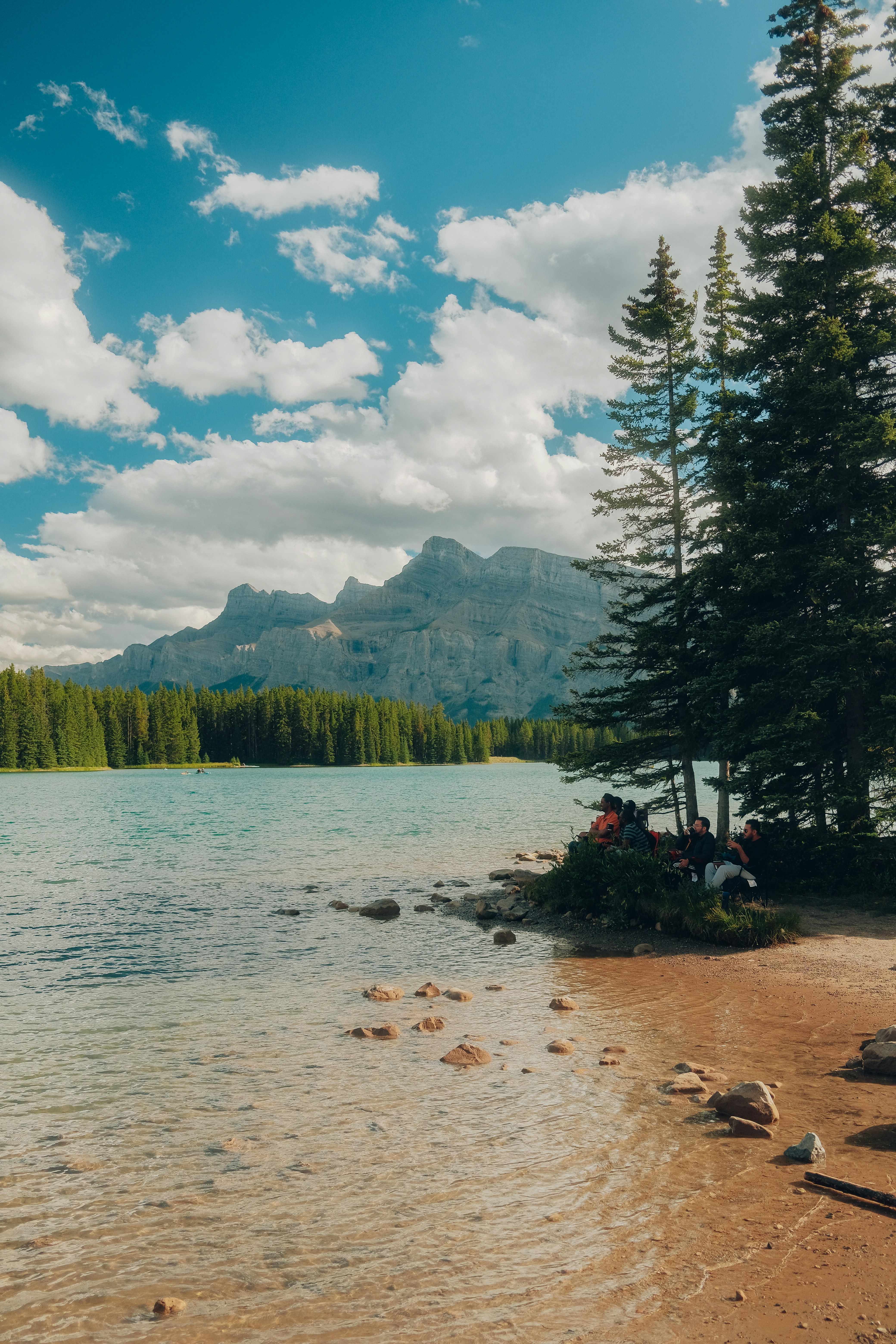 A serene lakeside scene featuring people gathered near the water's edge, framed by towering trees and majestic mountains under a cloudy sky.
