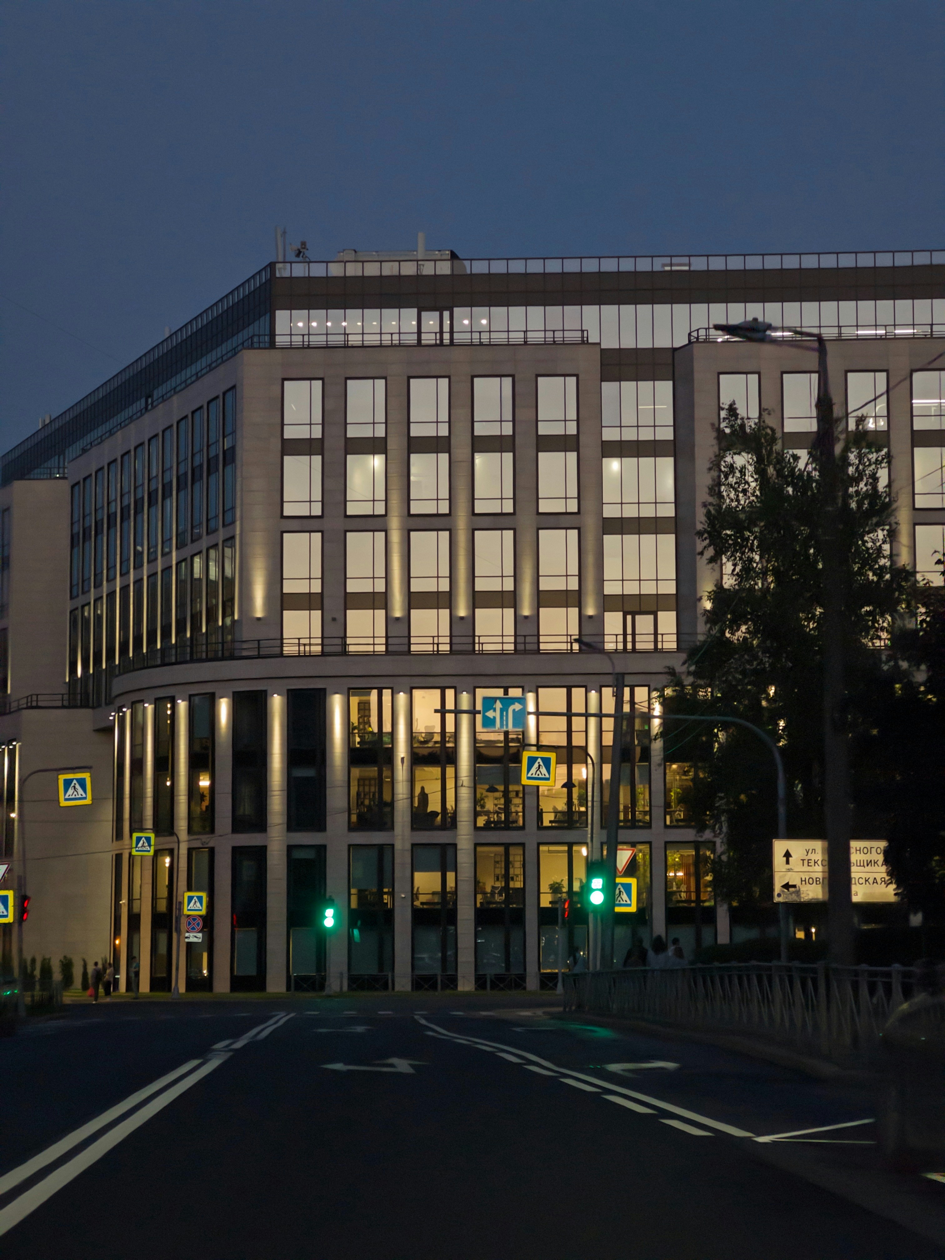 Illuminated office building reflecting twilight hues, framed by street signs and traffic lights.