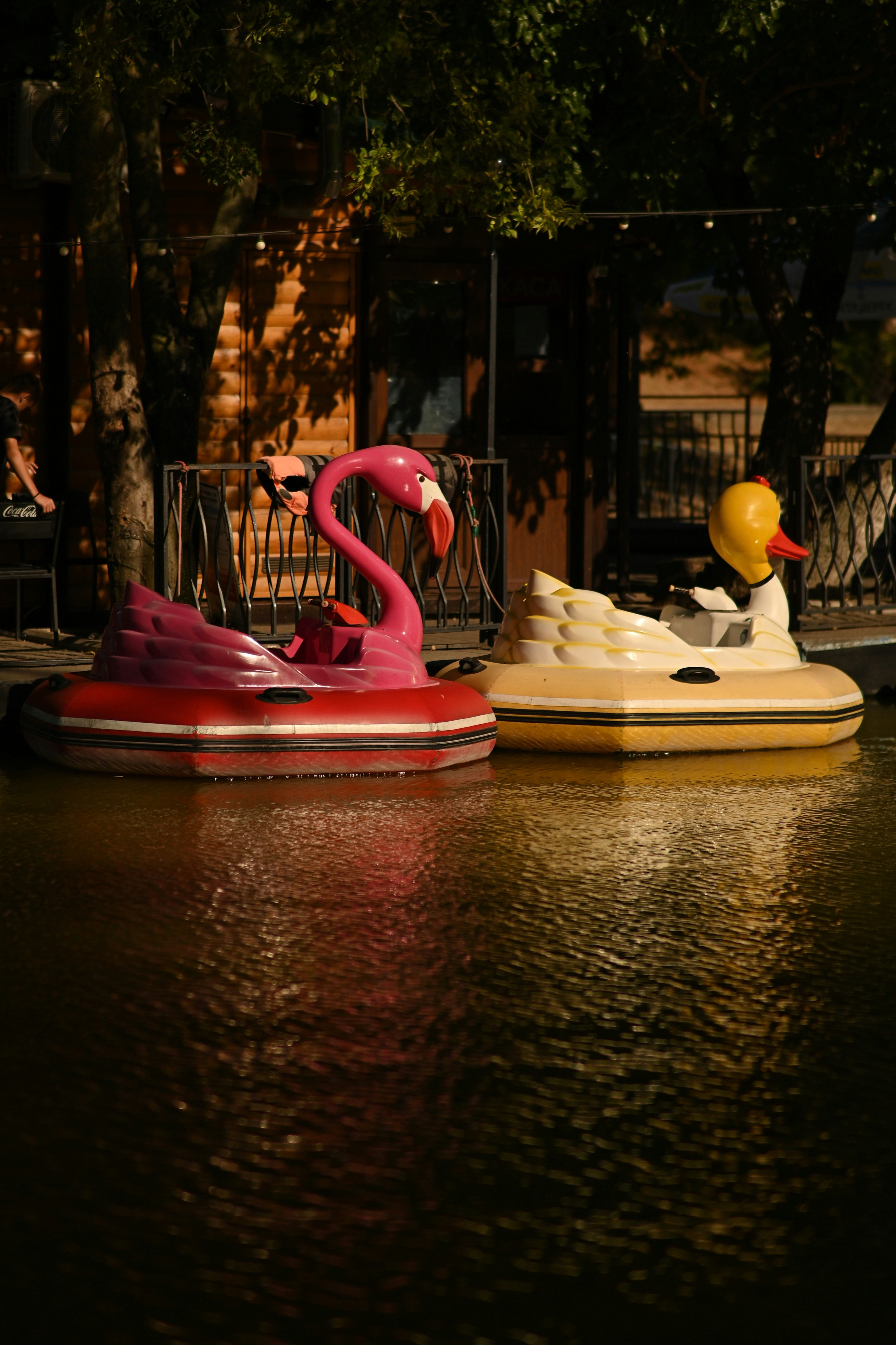 Two whimsical paddle boats, shaped like a pink flamingo and a yellow rubber duck, float side-by-side on a dark, rippling body of water. The warm light of the setting sun highlights their vibrant colors against the reflective surface. In the background, the scene is framed by the wooden facade of a building and the silhouettes of trees, creating a quiet and nostalgic atmosphere. | Two novelty pedal boats shaped like a flamingo and duck.