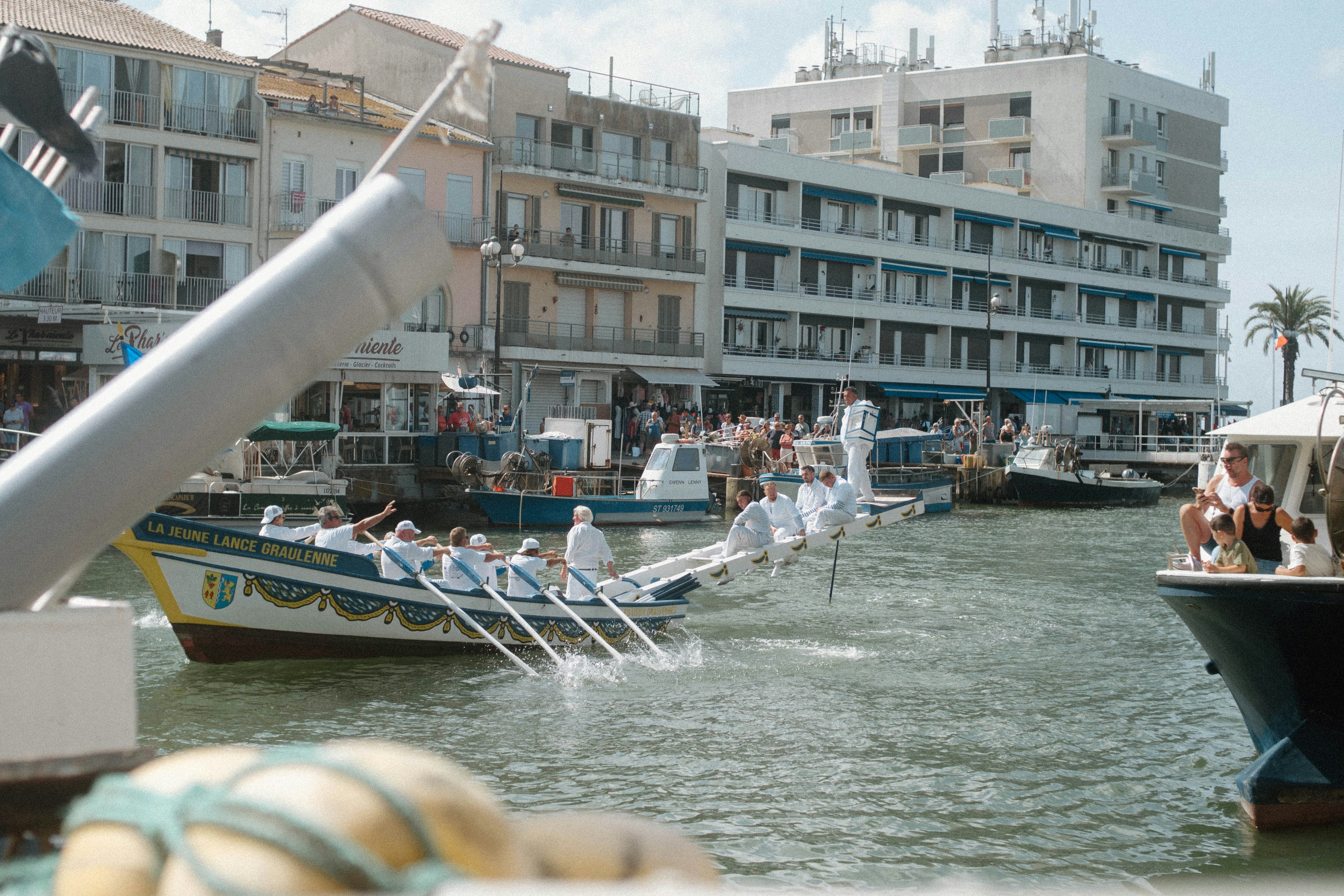 Rowers in a sunlit harbor sprint past a seaside town