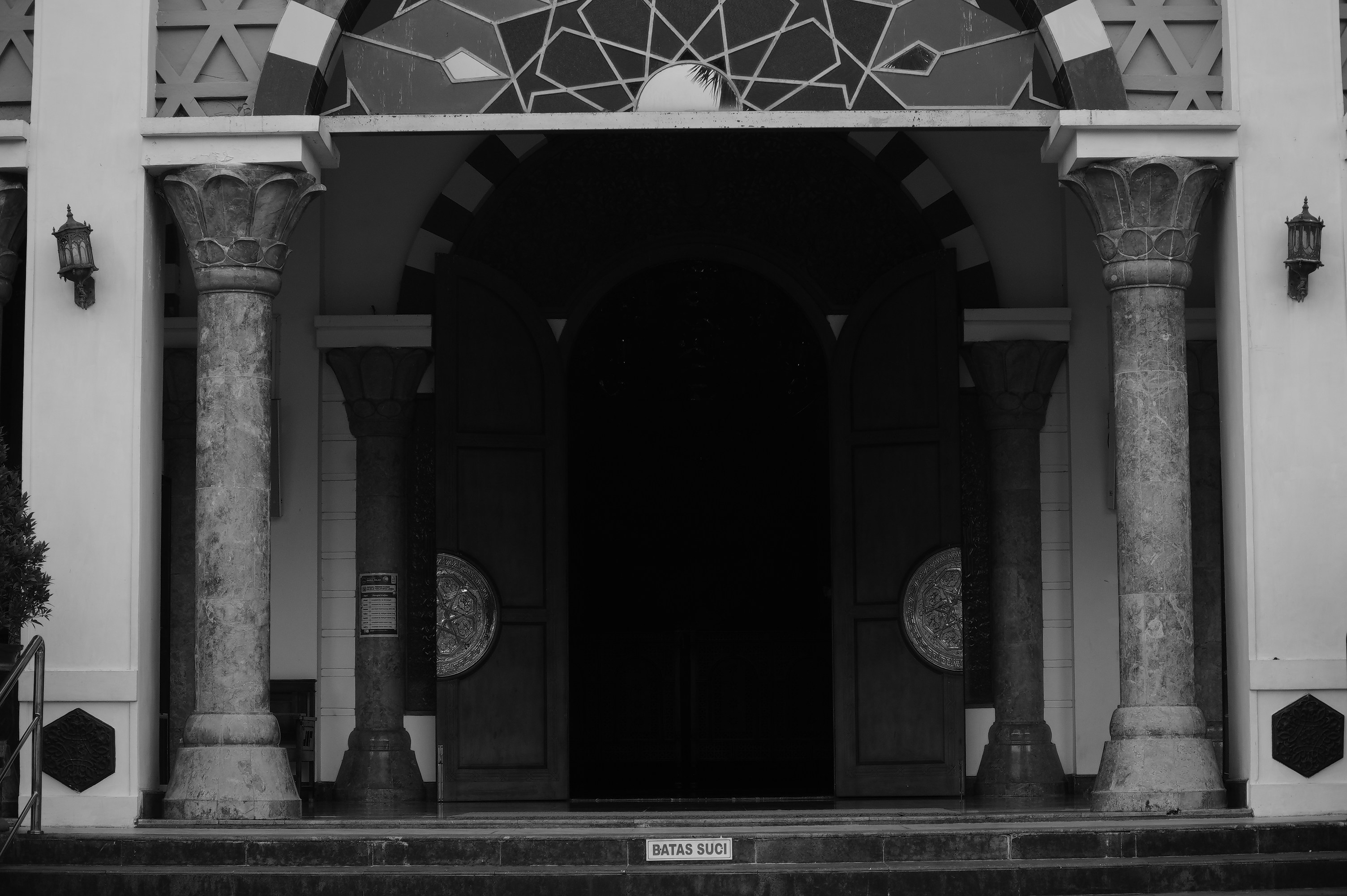 the entrance of a mosque, identified by the text overlay as Masjid Agung Darul Falah in Pacitan. The entrance features a large, dark arched doorway flanked by two prominent pillars with ornate capitals, resembling a classical architectural style. Above the entrance, there is decorative geometric patterning, typical of Islamic art. Two lamps are visible on either side of the entrance, mounted on the wall. The overall impression is one of a grand and architecturally significant religious building. | Entrance to a building with ornate columns and archway.