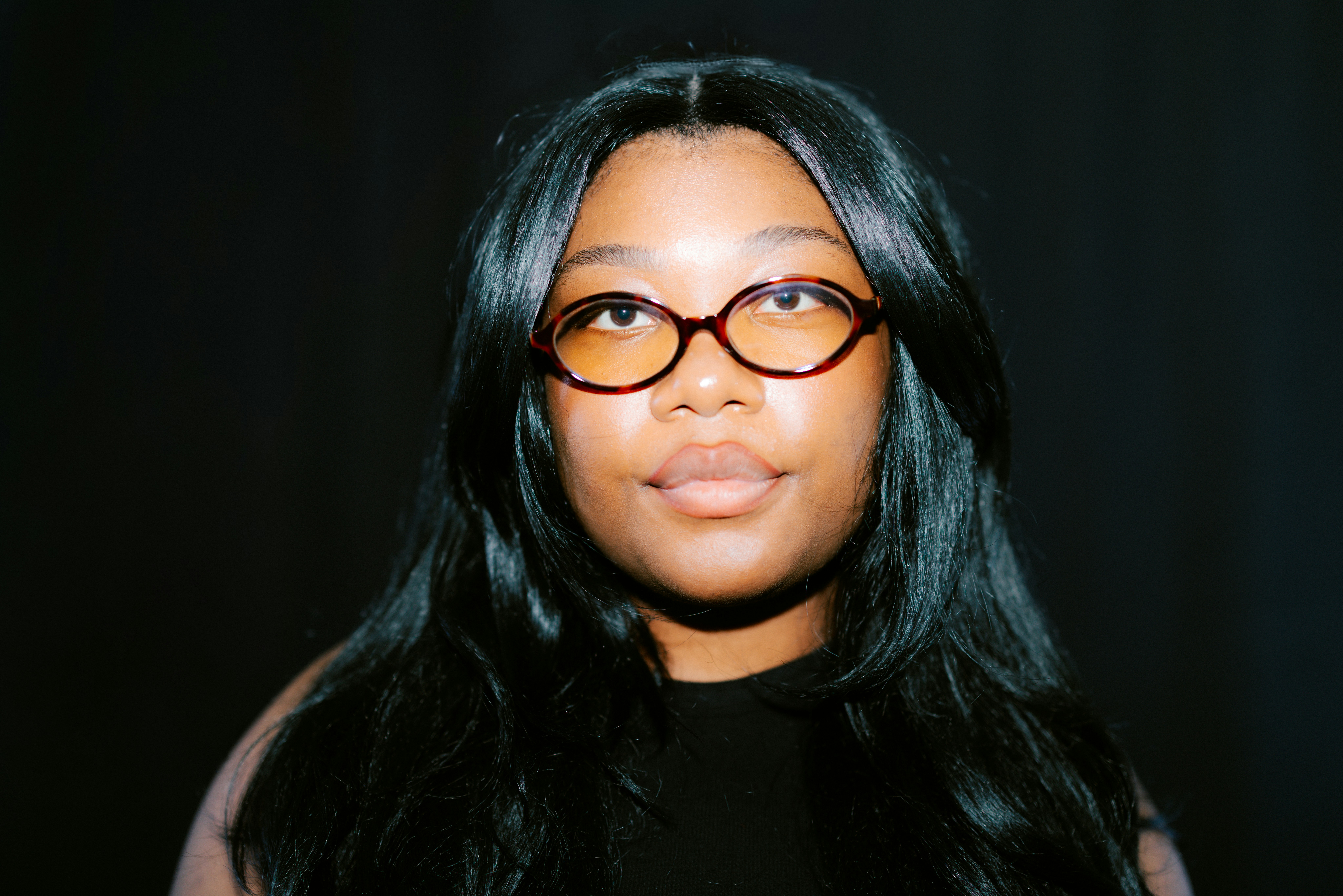 A young woman with long, dark hair and stylish glasses gazes thoughtfully against a dark backdrop.