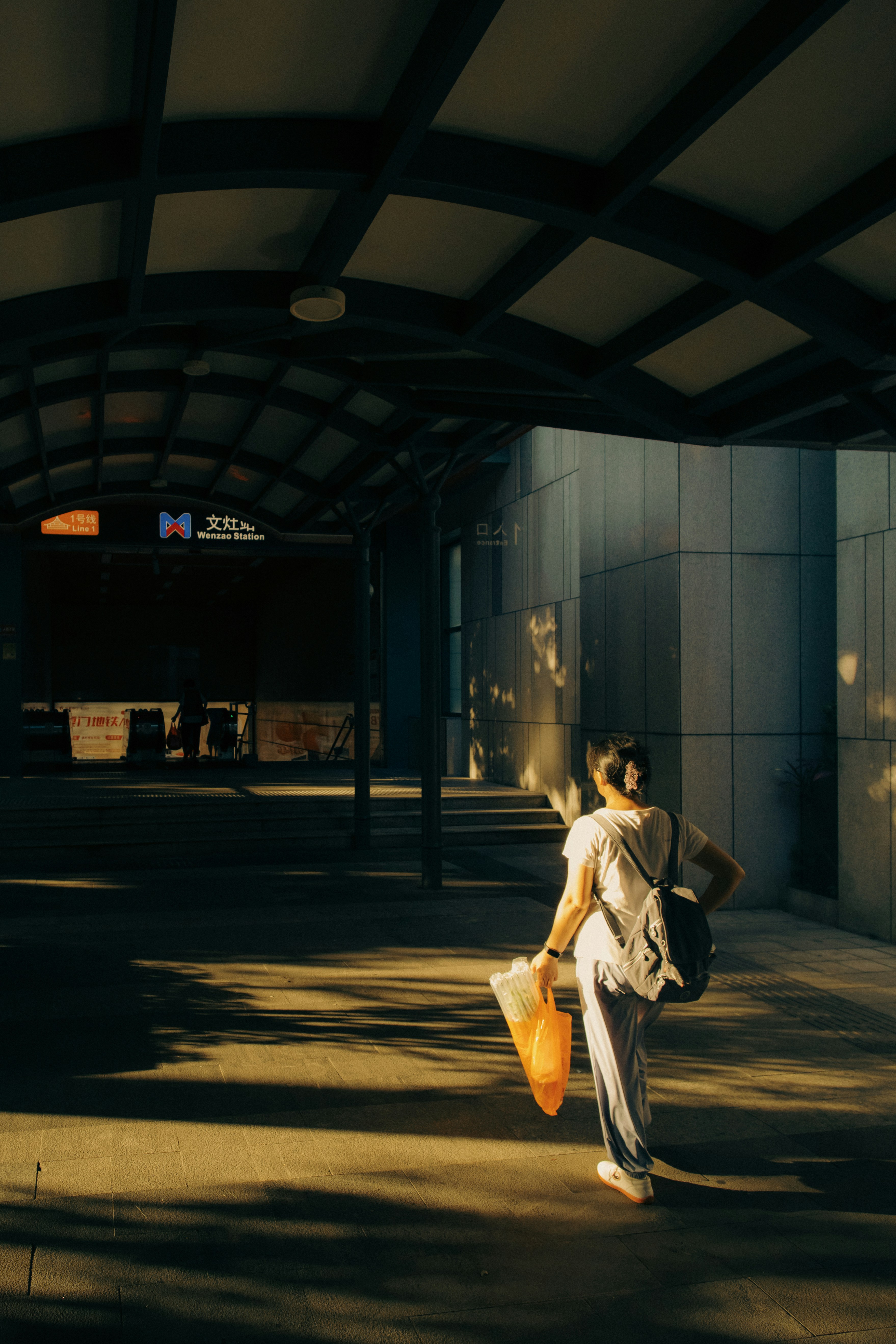 A person walks through a sunlit urban corridor, carrying an orange bag, with shadows creating a dramatic interplay of light and architecture.
