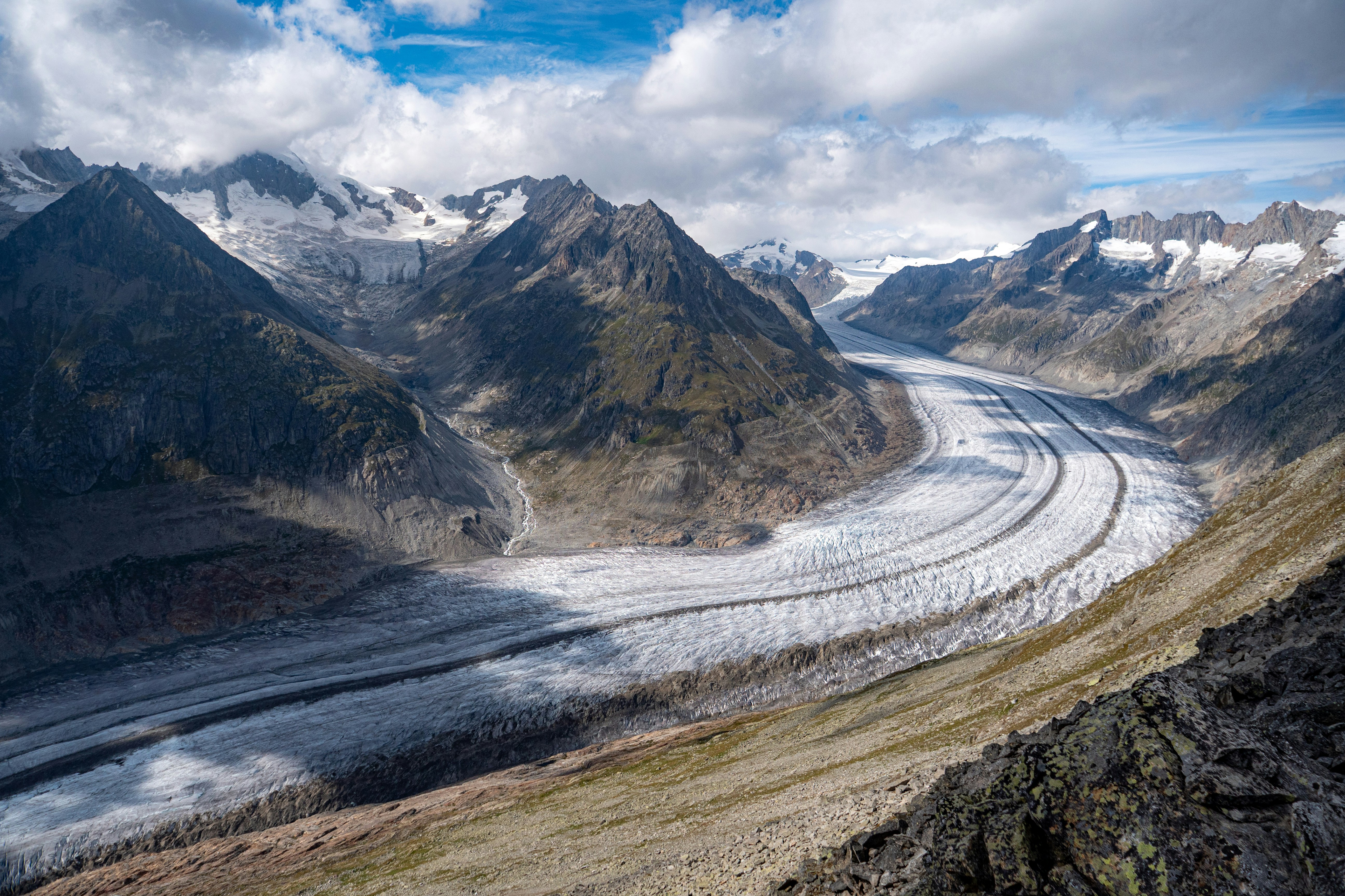 A sweeping view of a glacier winding through majestic mountains under a partly cloudy sky. The glacial patterns create a striking contrast against the rugged terrain.
