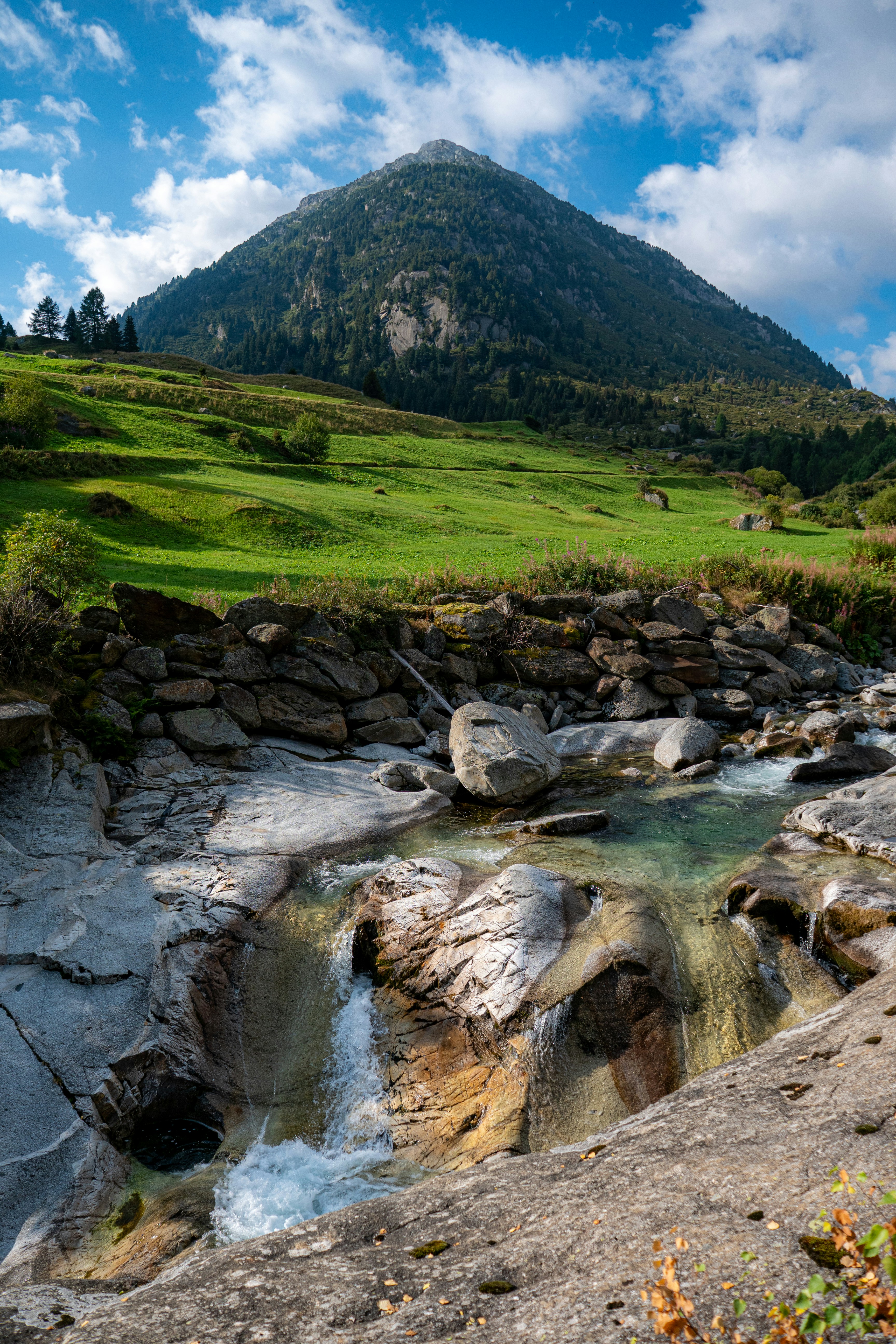 Gentle stream cascading over smooth rocks in a lush valley, framed by a towering mountain under a partly cloudy sky.