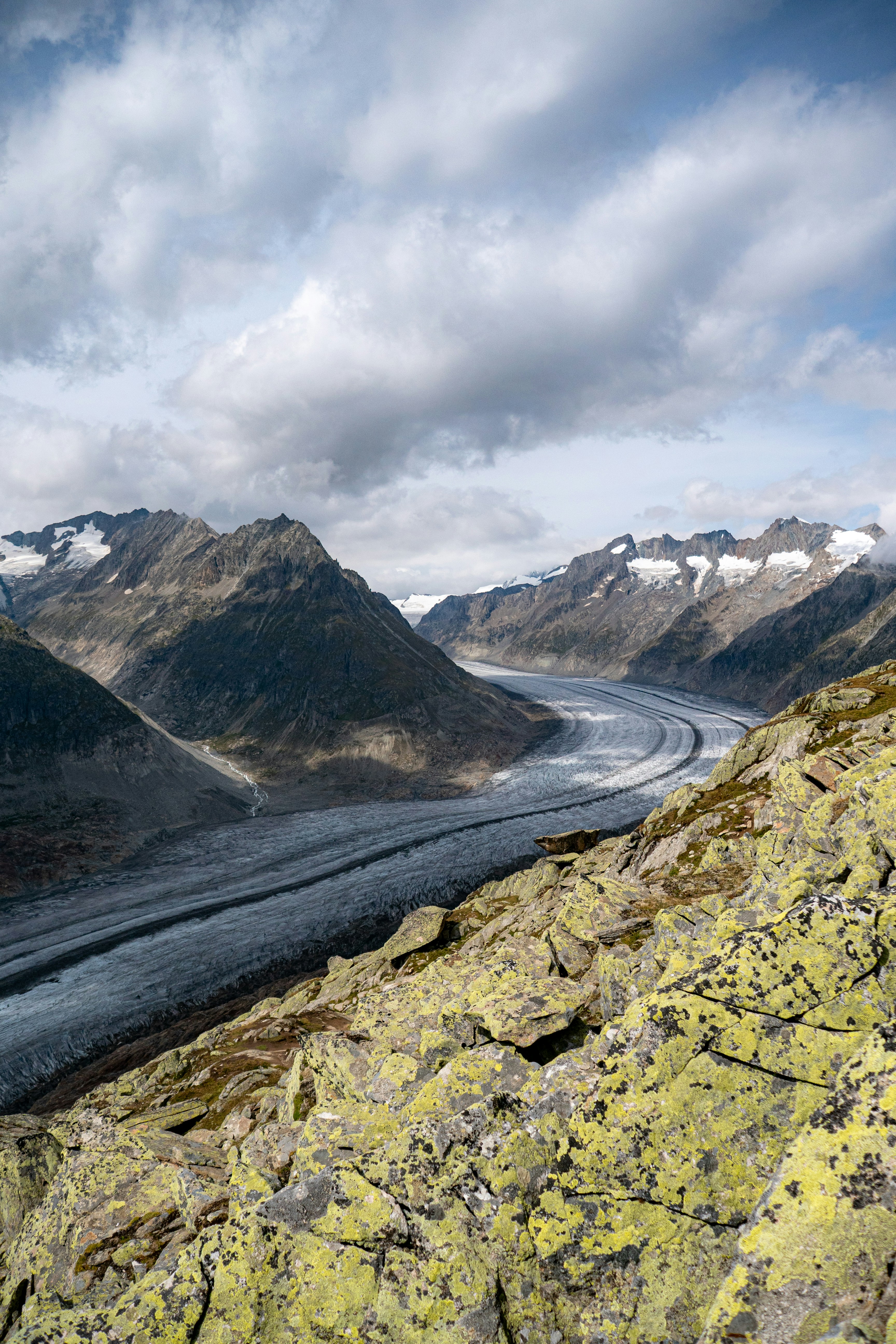 Winding glacier flows gracefully between towering mountains under a dynamic sky, showcasing nature's grandeur. The rugged foreground features moss-covered rocks.