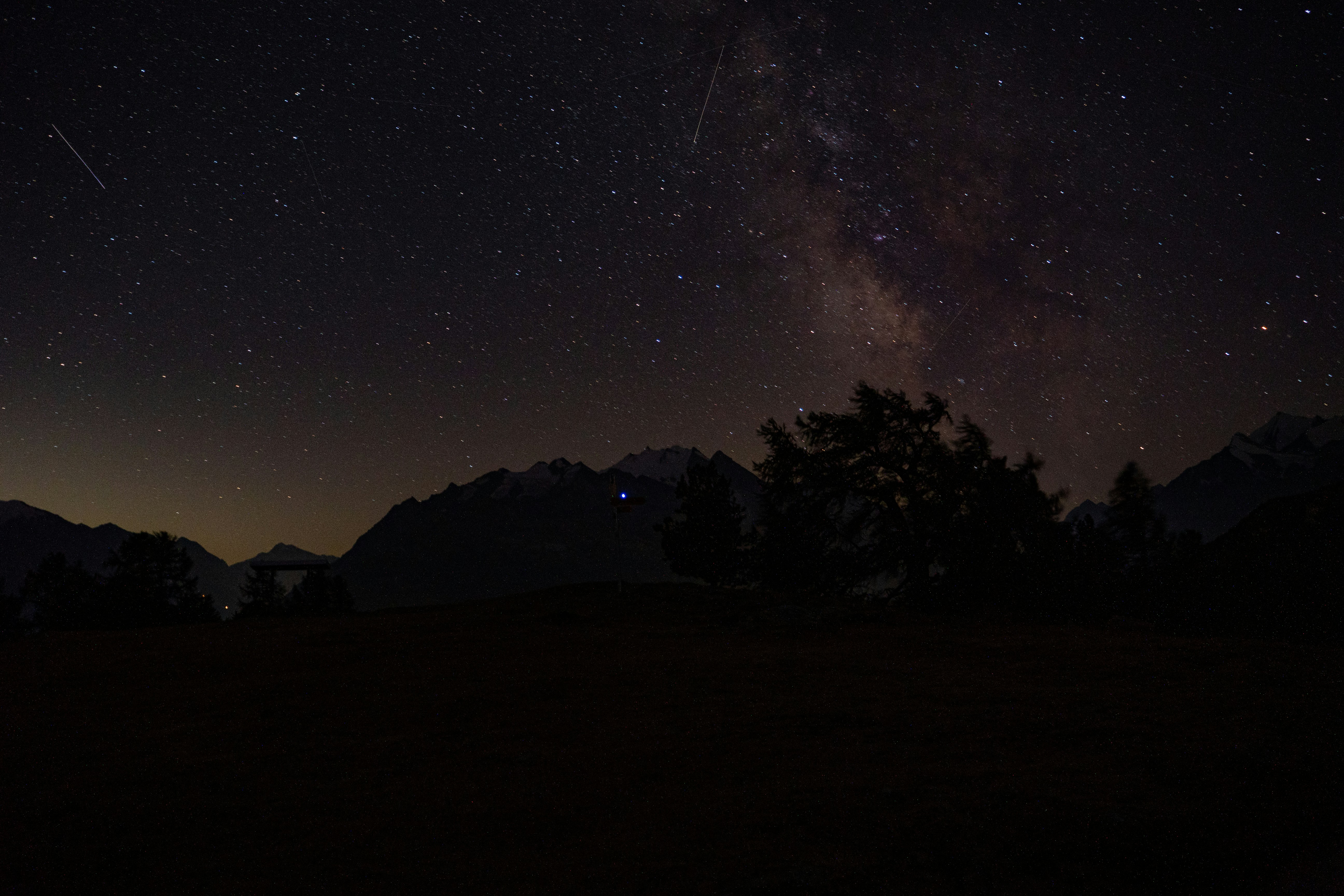 Starry night sky over mountains with faint trees.