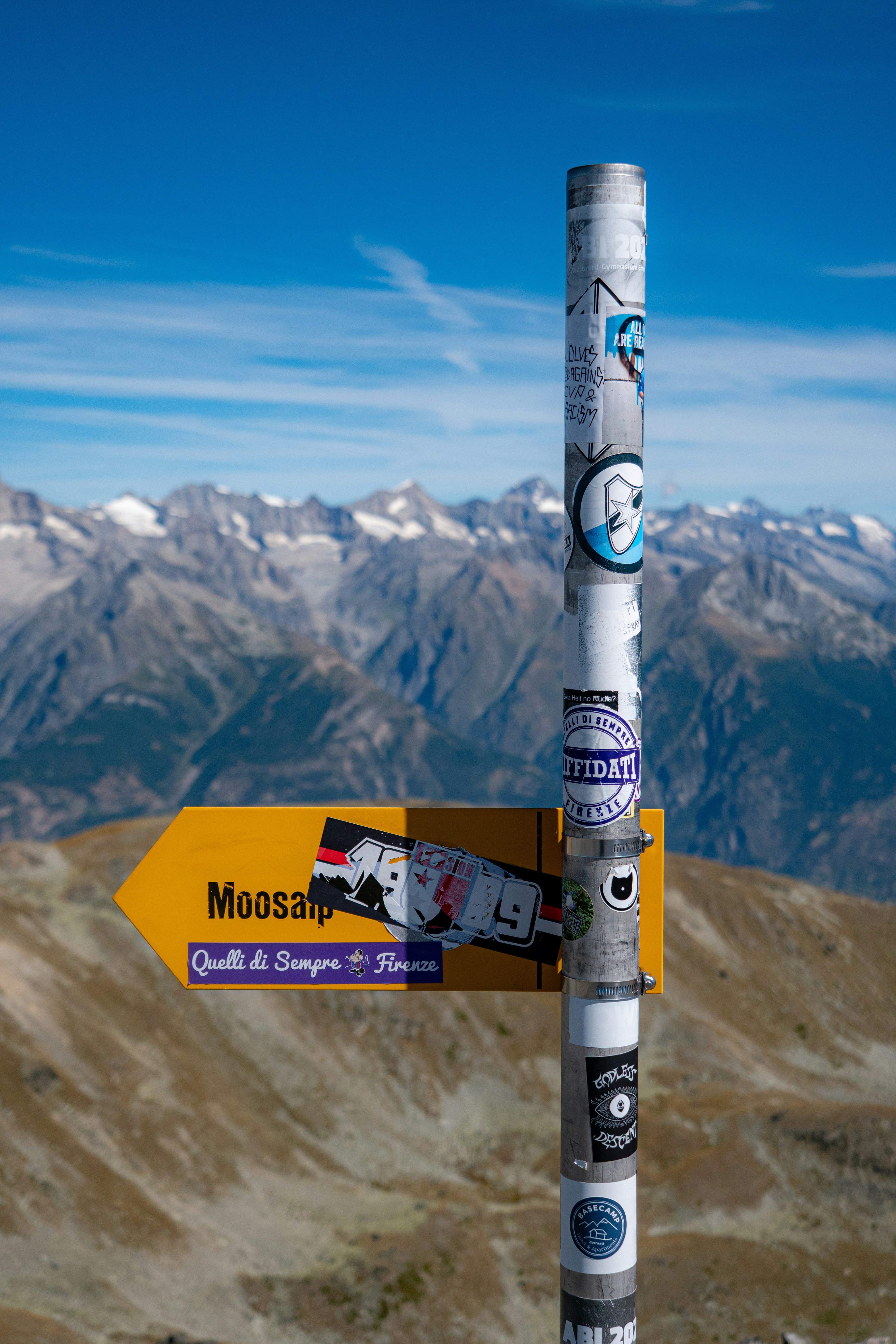 Directional signpost adorned with stickers, overlooking a breathtaking mountain landscape. The sign points towards Moosalp, inviting adventurers to explore the scenic routes.