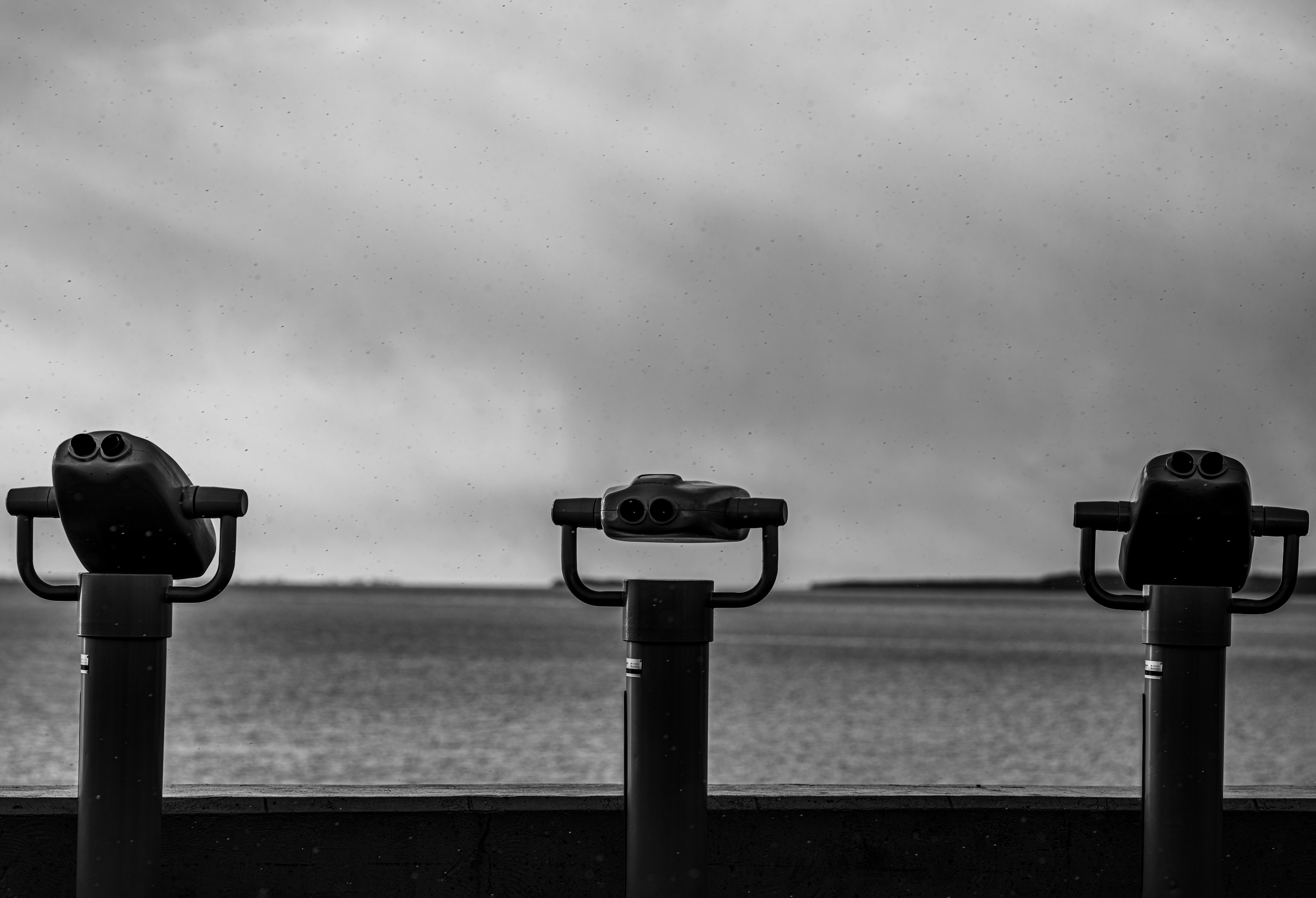 Three binoculars stand on a railing, overlooking a calm body of water under a moody sky.