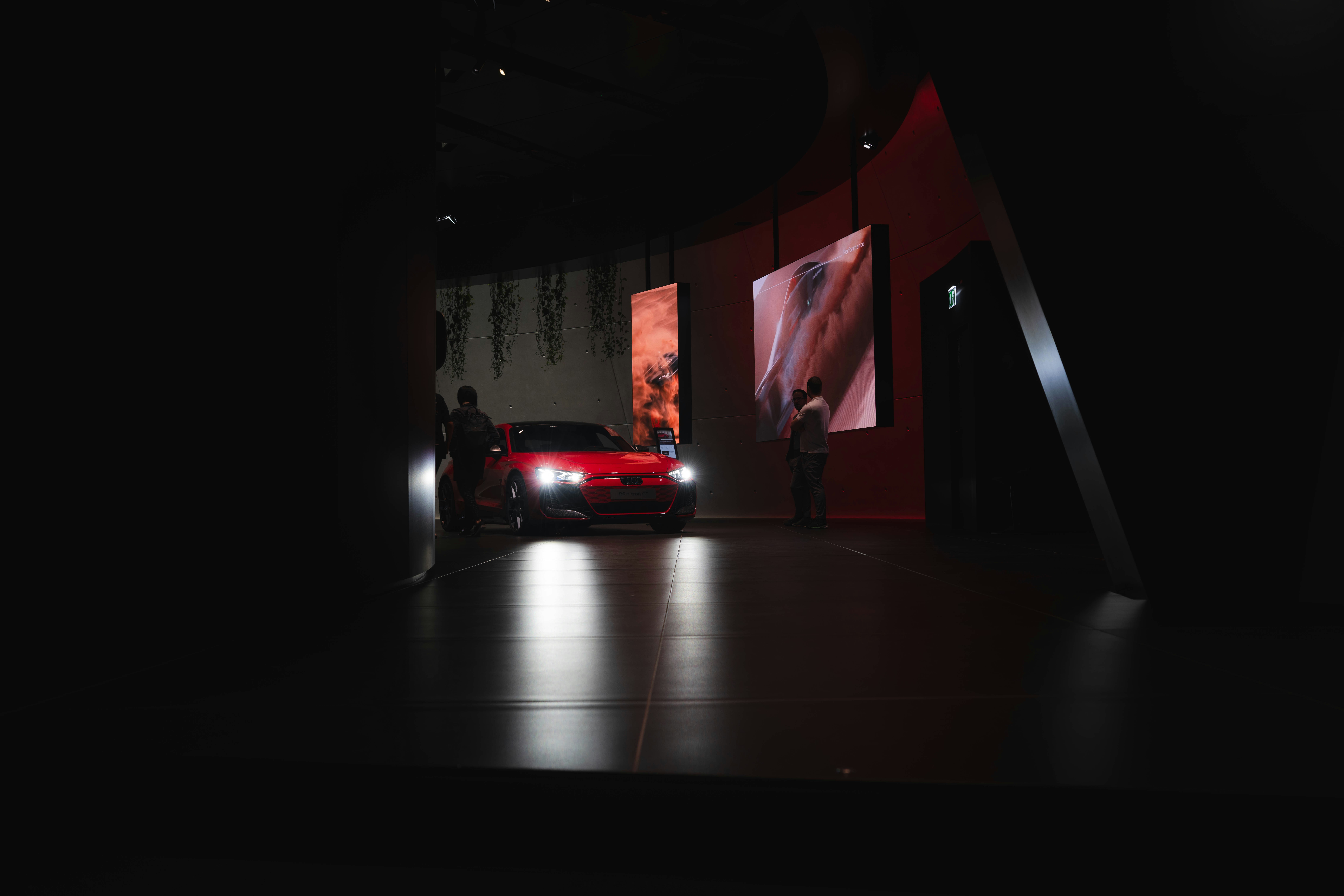 A sleek red car is showcased in a dimly lit space, highlighting its design against vibrant digital displays. The atmosphere is enhanced by dramatic lighting.
