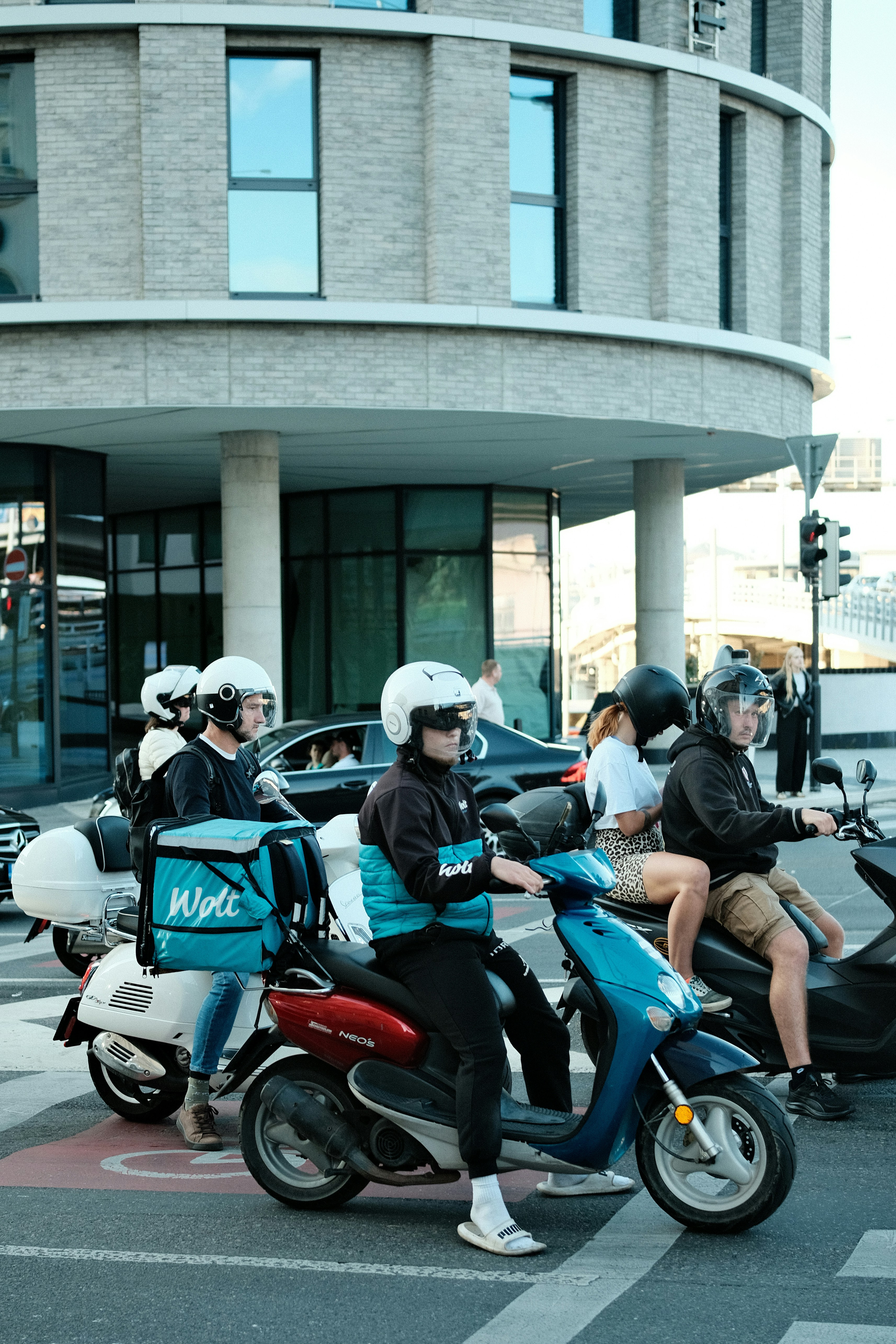 Scooter delivery workers waiting at traffic light in urban setting | Delivery drivers on scooters wait at intersection