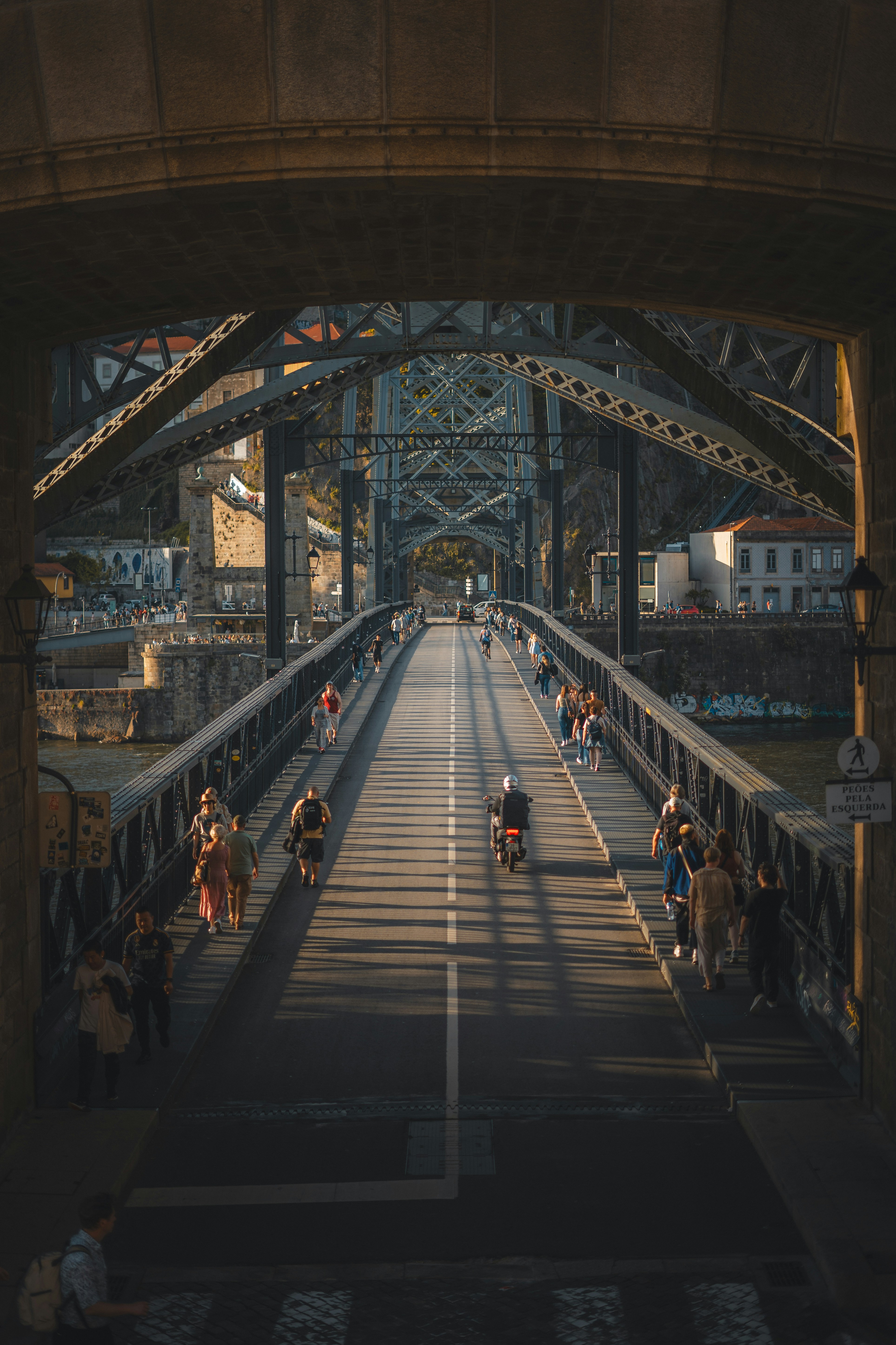 People walking and cycling across a bridge, framed by intricate steel architecture and warm evening light.