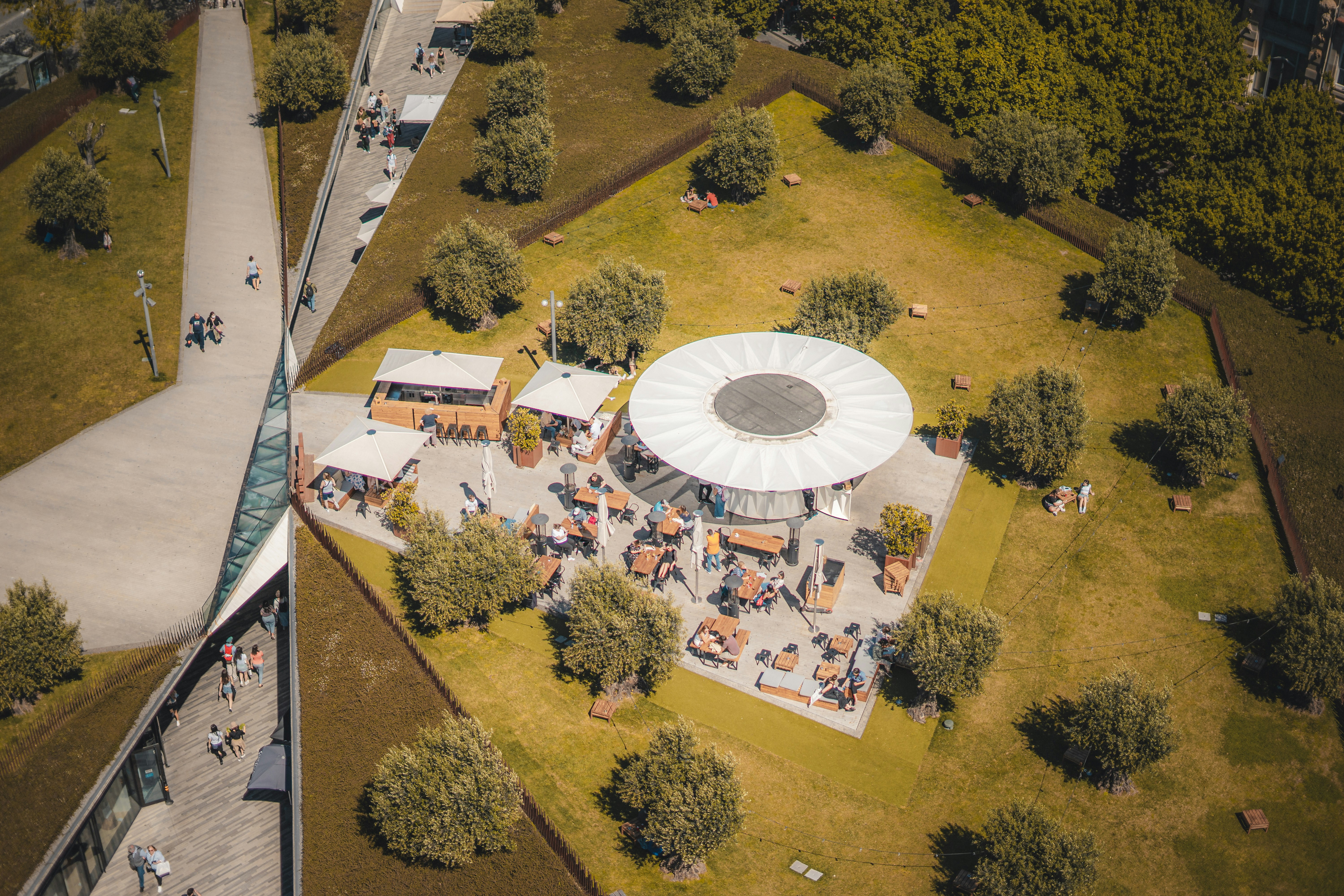 A vibrant outdoor café surrounded by lush greenery, featuring circular seating and white canopies, bustling with visitors enjoying the sunny day.