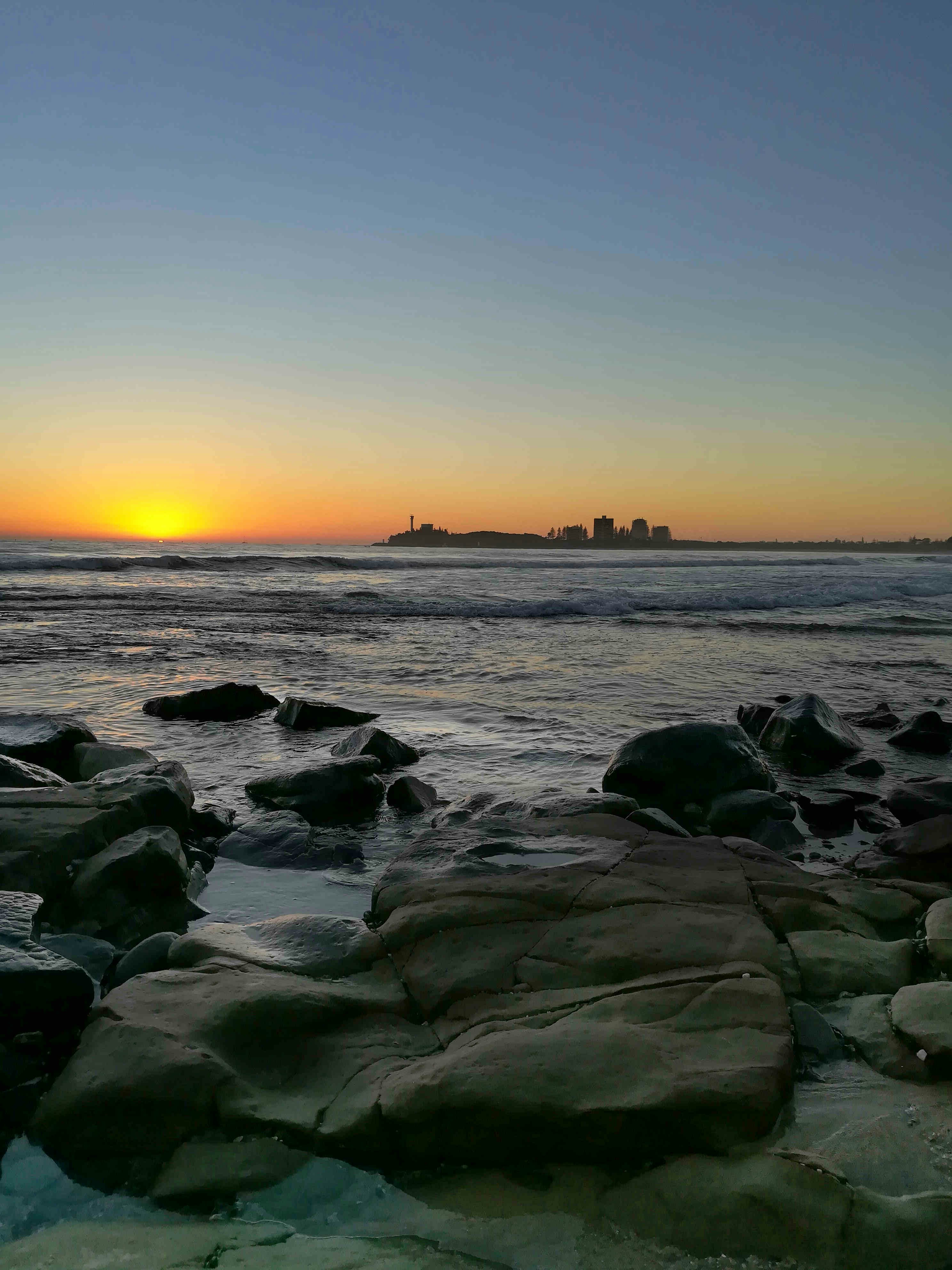 Sunset over ocean with rocky shore and city skyline.