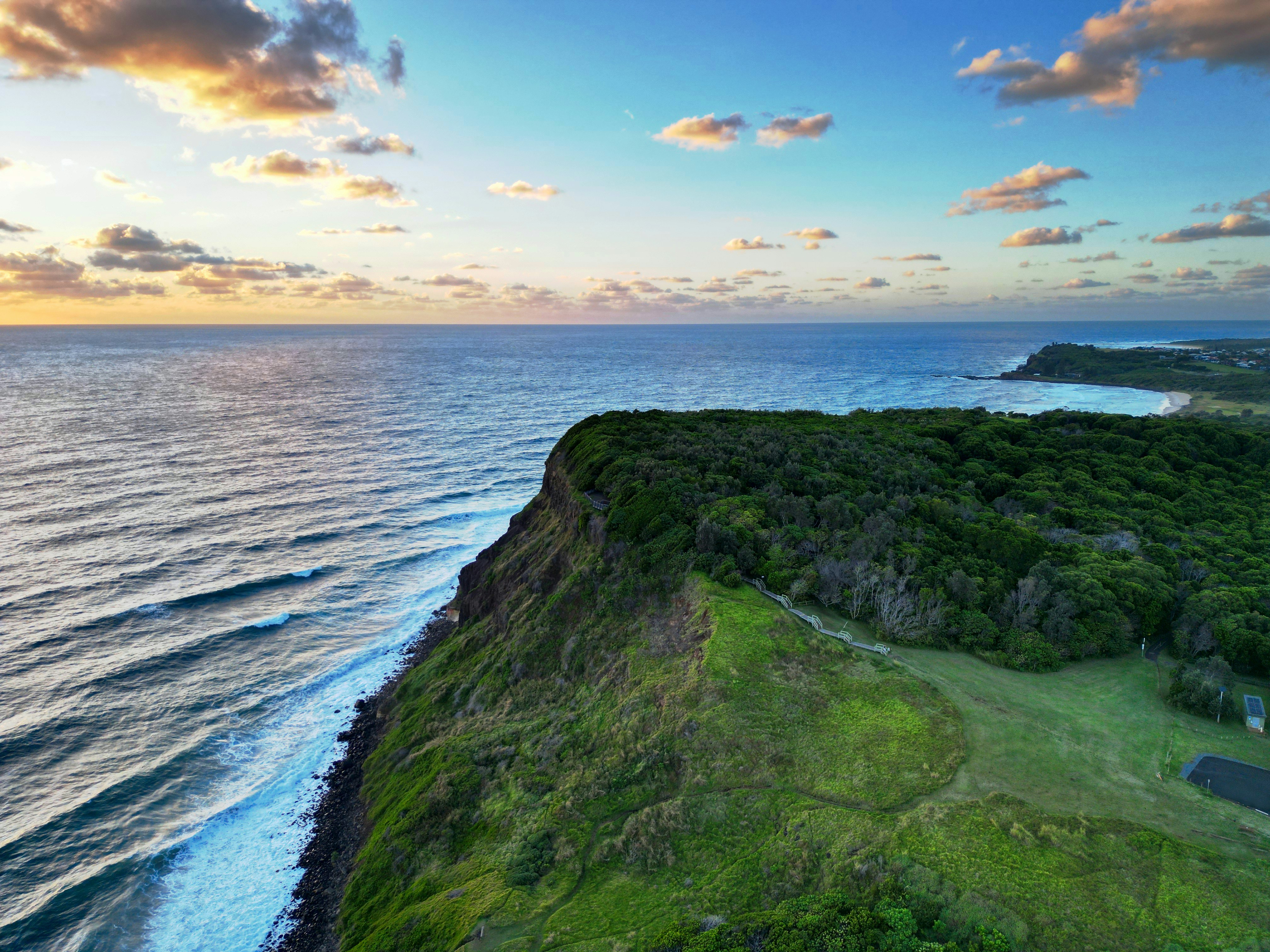 Aerial view of a lush green cliffside overlooking the ocean at sunrise, with gentle waves lapping against the shore.