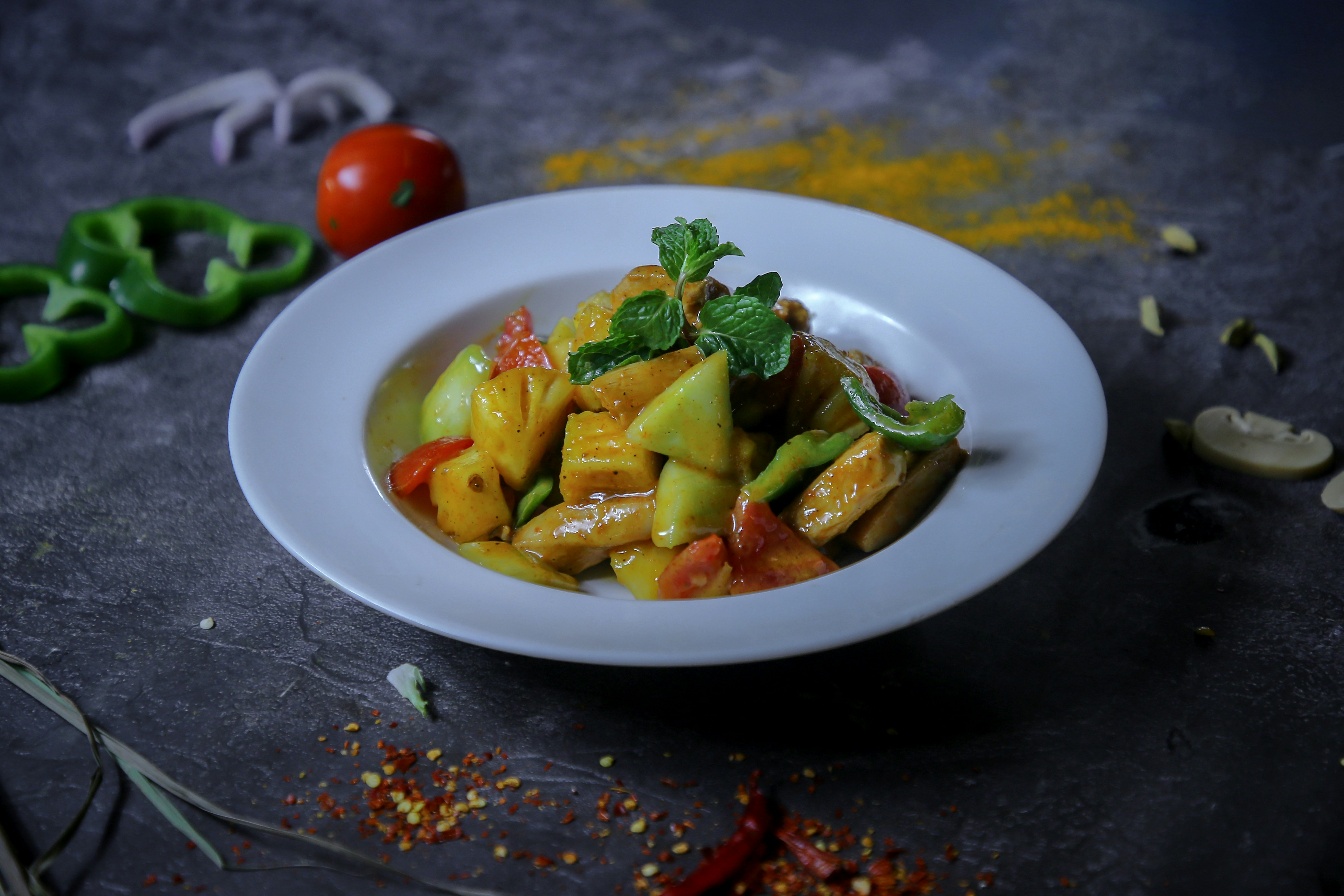 A close-up of a stir-fried dish on a white plate. It contains chicken and vegetables, with a glossy sauce and a mint garnish. Spices and fresh ingredients surround the plate. | A delicious dish of paneer tikka with fresh mint leaves.