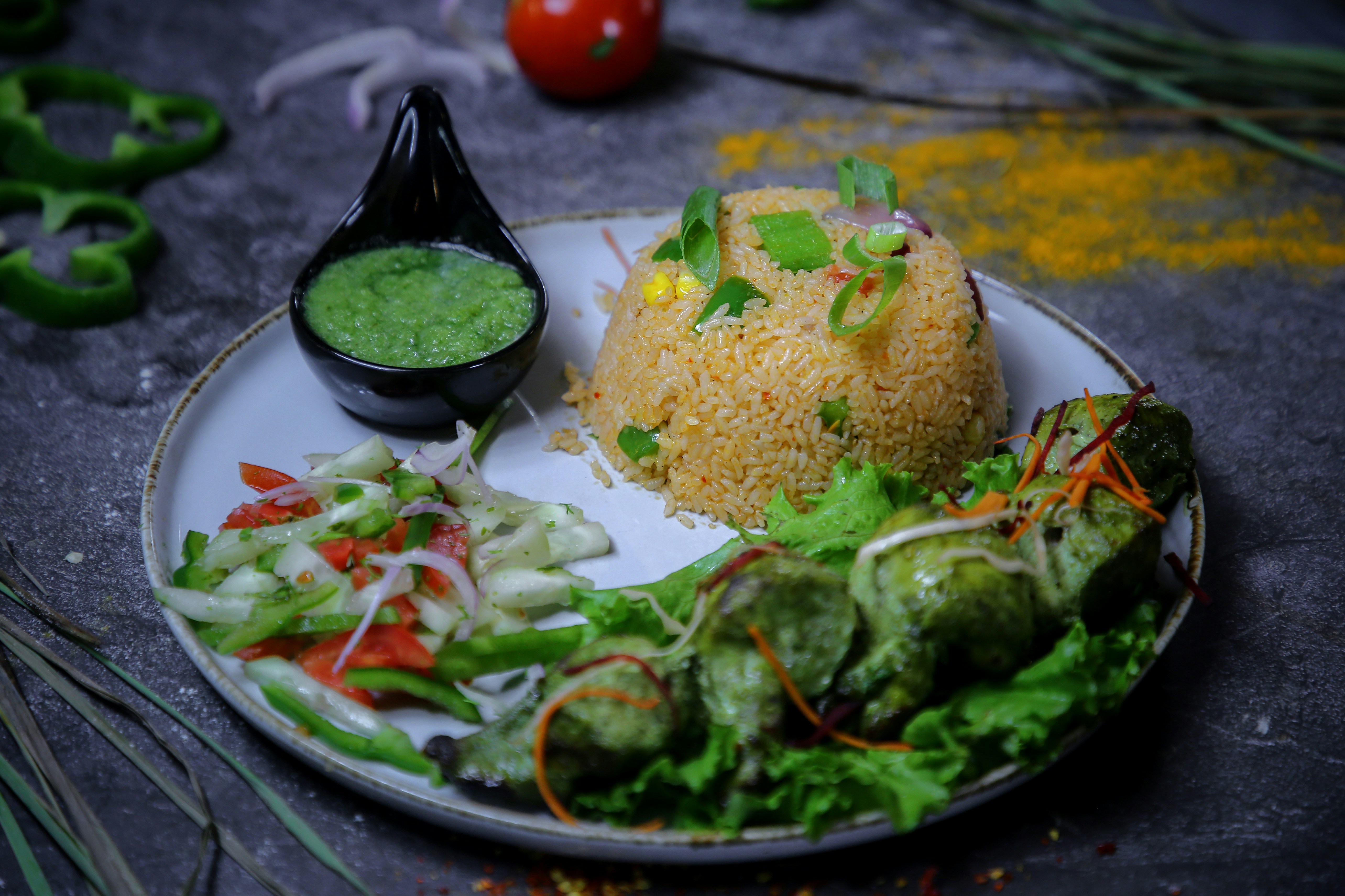 A delicious meal served on a modern plate. It features a mound of fried rice, alongside juicy green-marinated chicken pieces on a bed of lettuce. A side of fresh salad and a dipping sauce completes the vibrant and appetizing dish. | Green chicken tikka with rice and salad