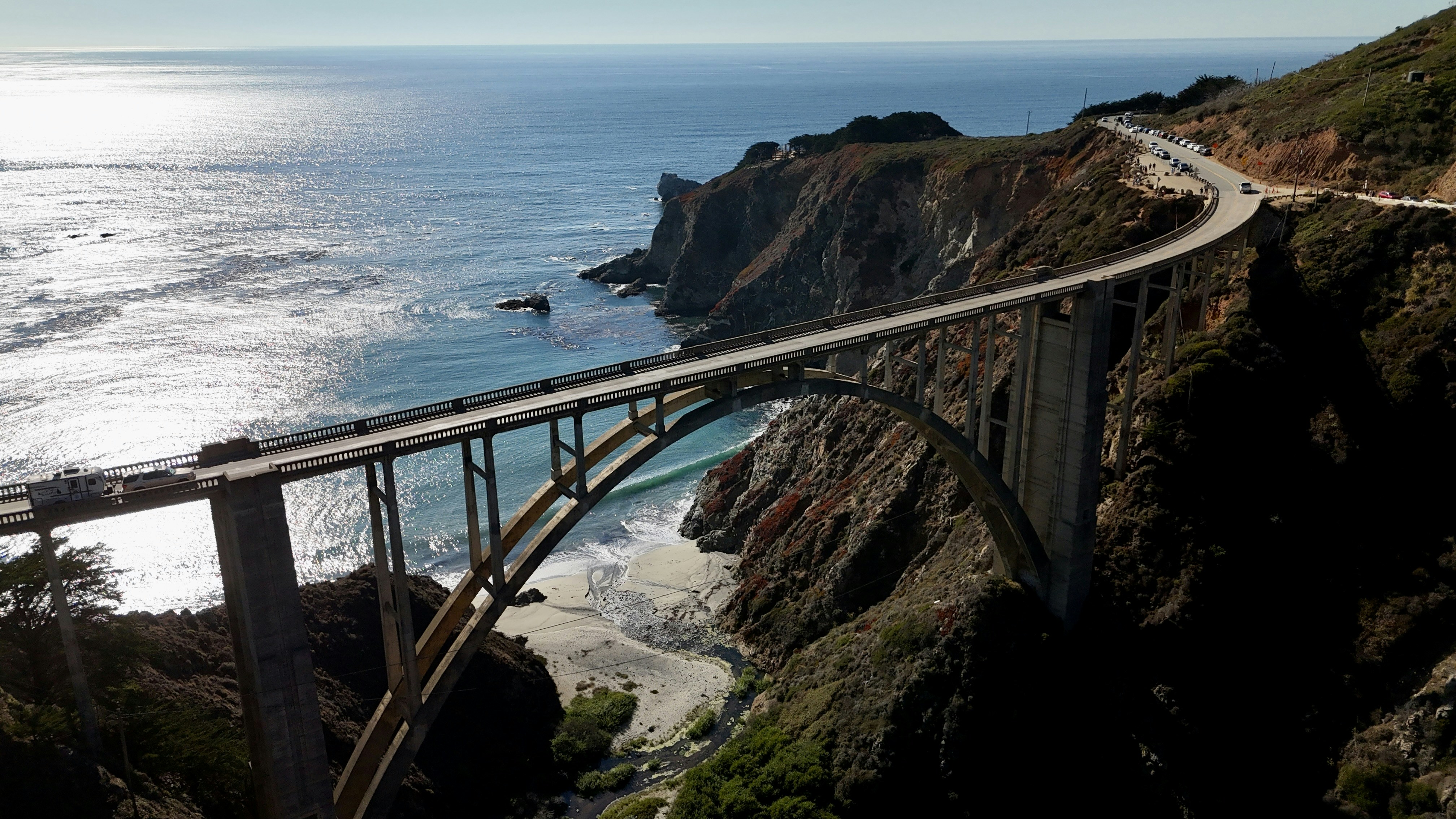 The iconic Bixby Creek Bridge stretches across a rugged cliffside along California’s Highway 1 in Big Sur, overlooking the shimmering Pacific Ocean. Its dramatic concrete arch rises above a secluded beach and rocky coastline, blending engineering brilliance with breathtaking natural scenery. This world-famous landmark is a highlight of the Pacific Coast Highway and a must-see stop for travelers and photographers. | Bixby creek bridge on a sunny day