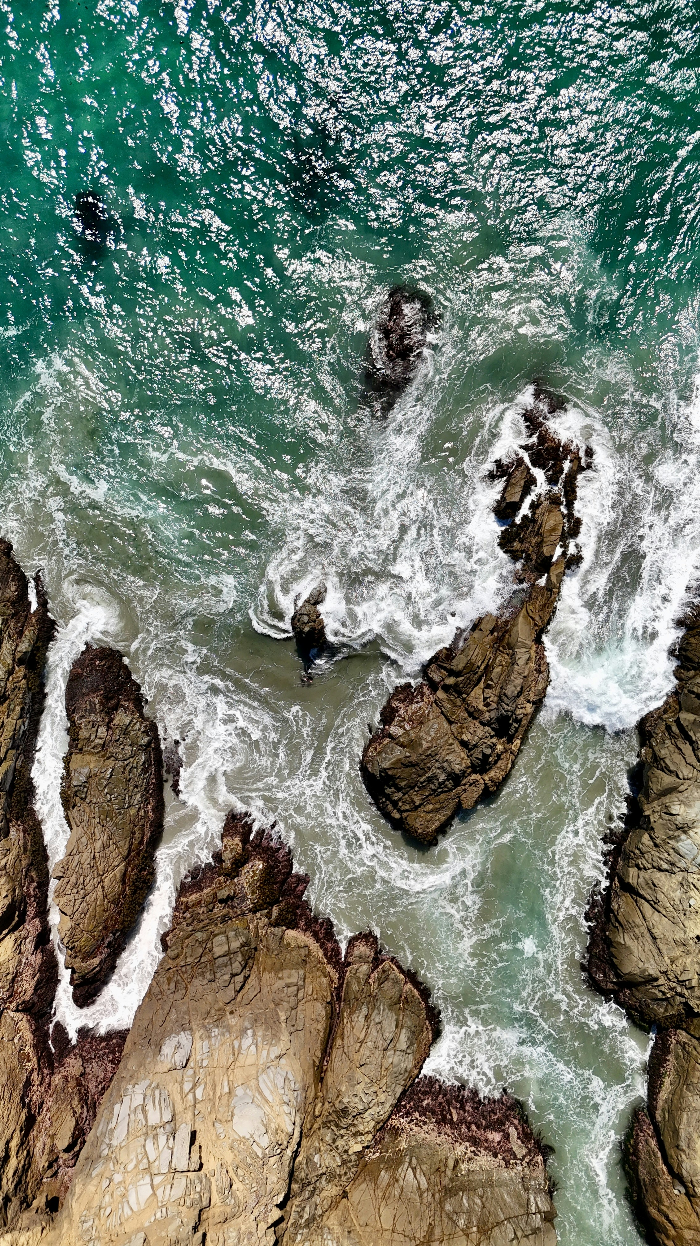 Aerial view showcasing rugged rocks partially submerged in vibrant turquoise waters, with waves crashing against the shoreline.