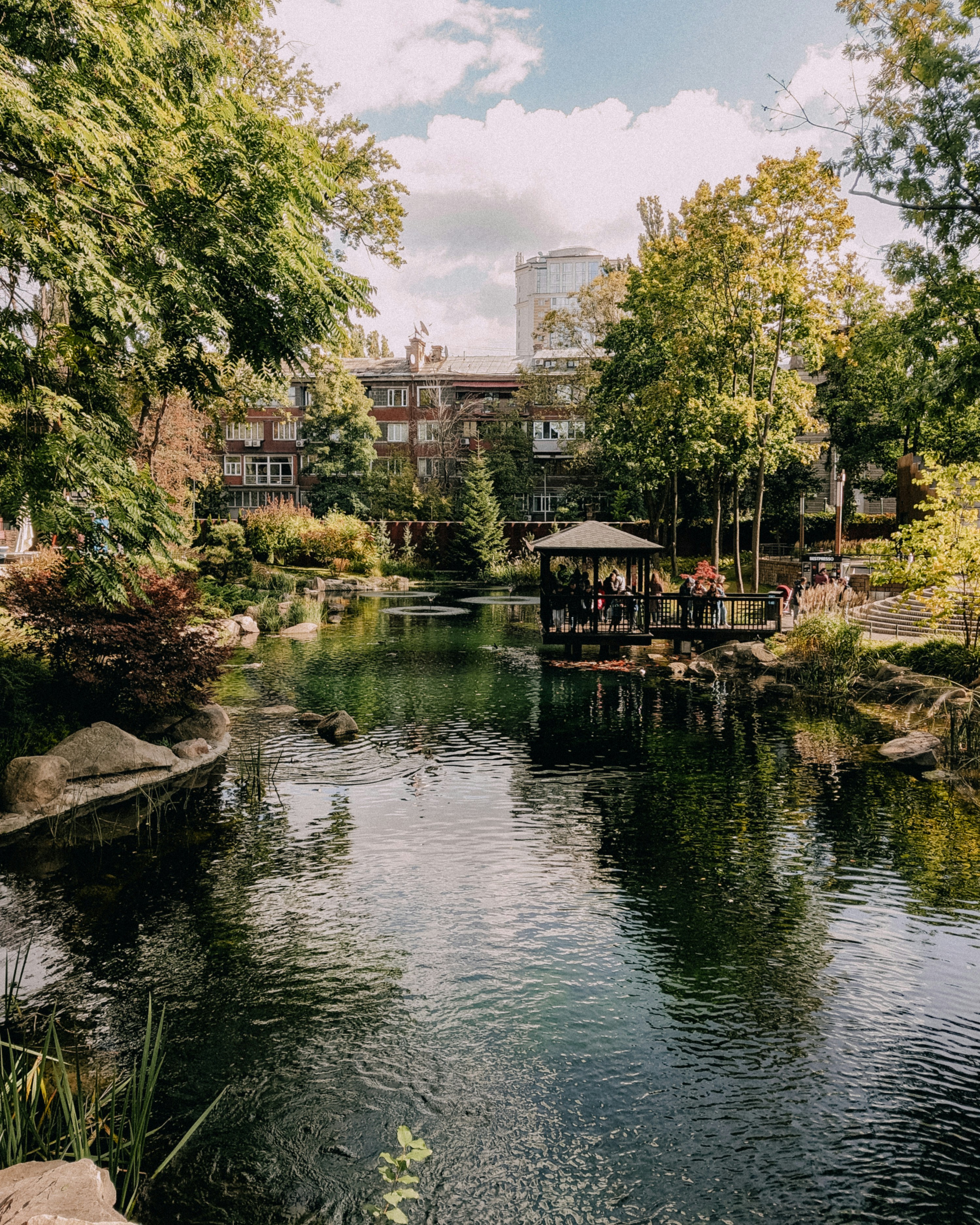 Tranquil park scene featuring a pond surrounded by lush trees and a gazebo, with people enjoying the serene atmosphere.