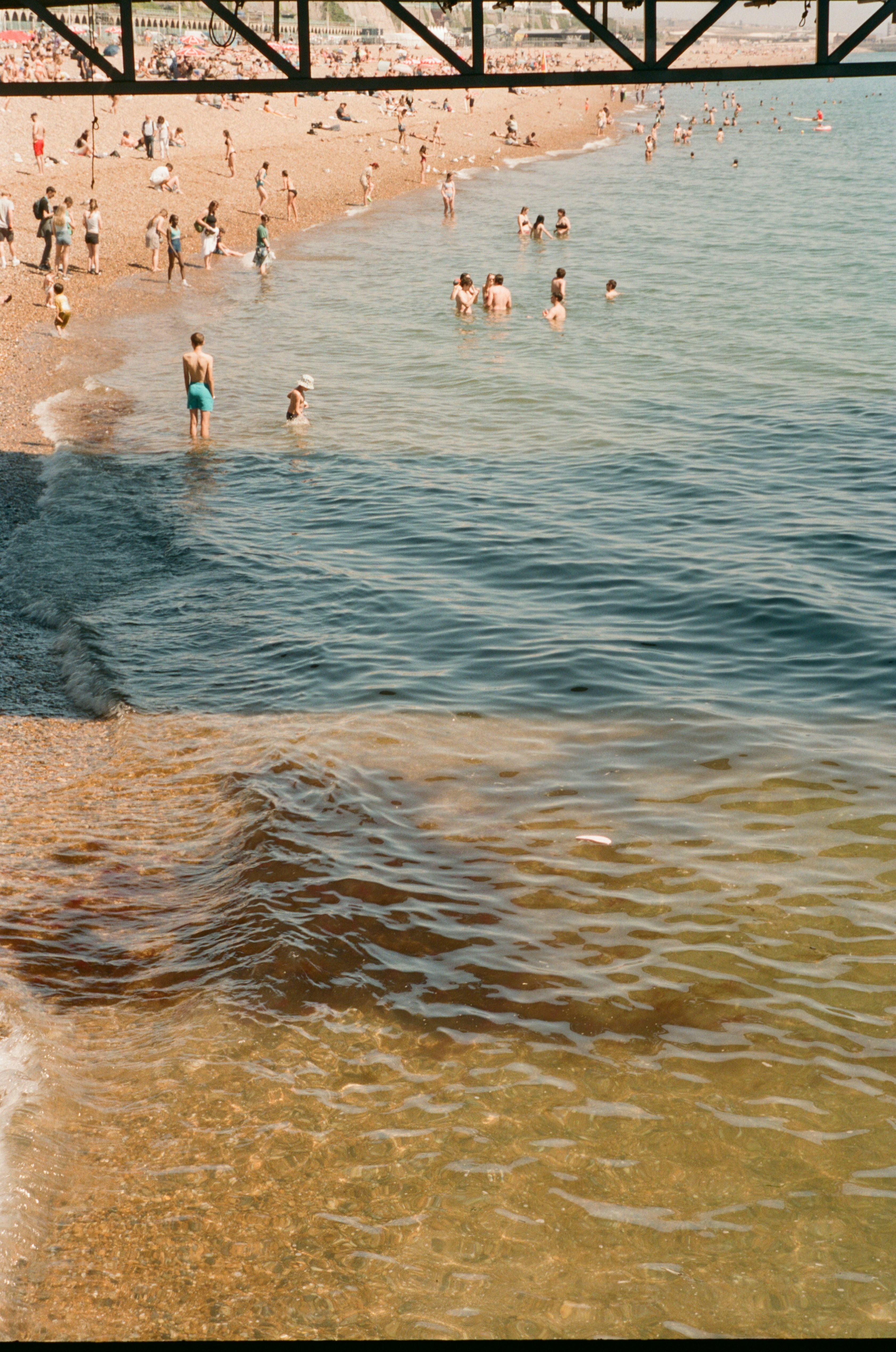Beachgoers enjoying a sunny day by the water under a pier, with gentle waves lapping at the shore.