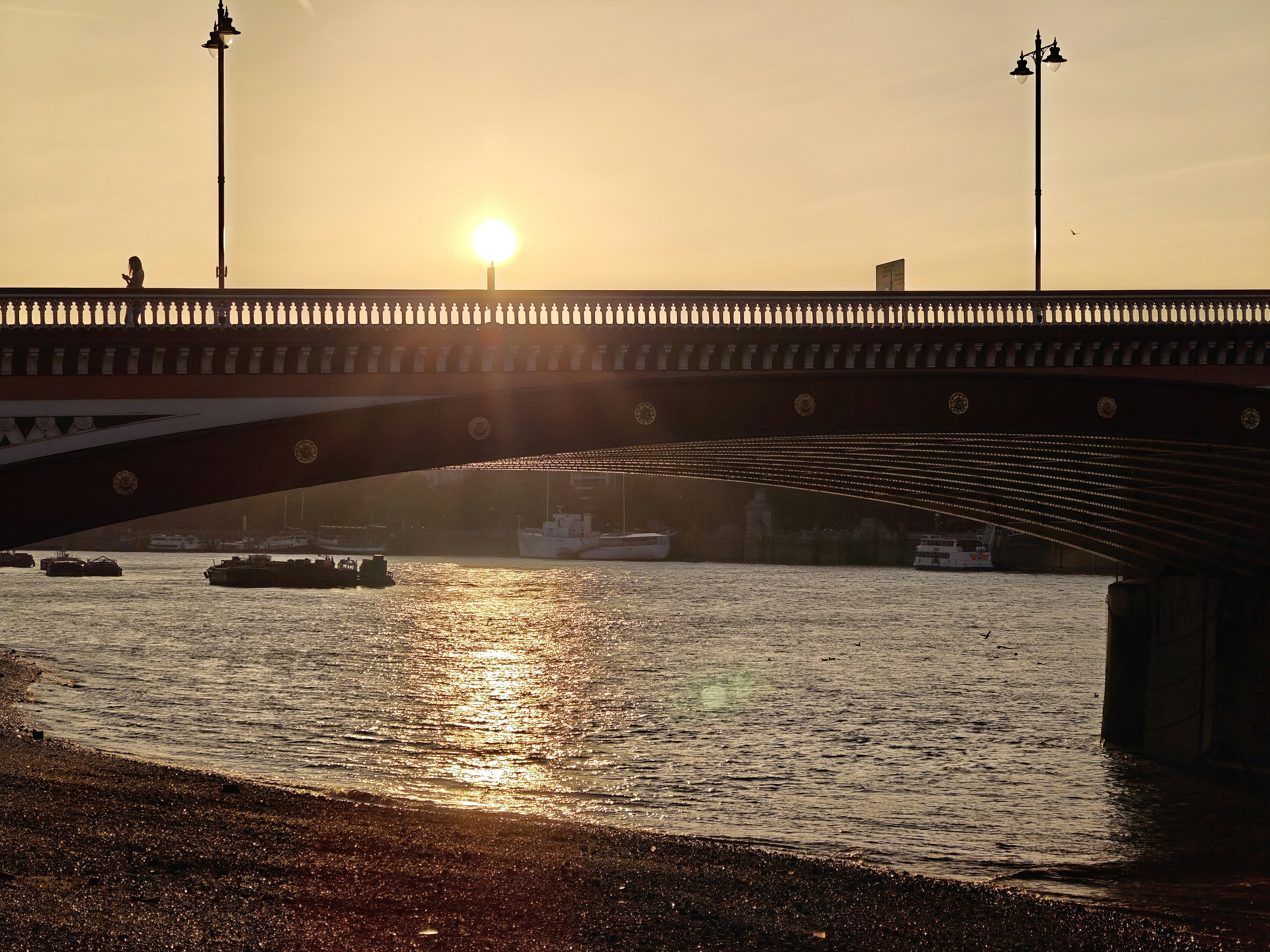 Silhouetted figure walks along a bridge at sunset, casting a long shadow over the shimmering river below.