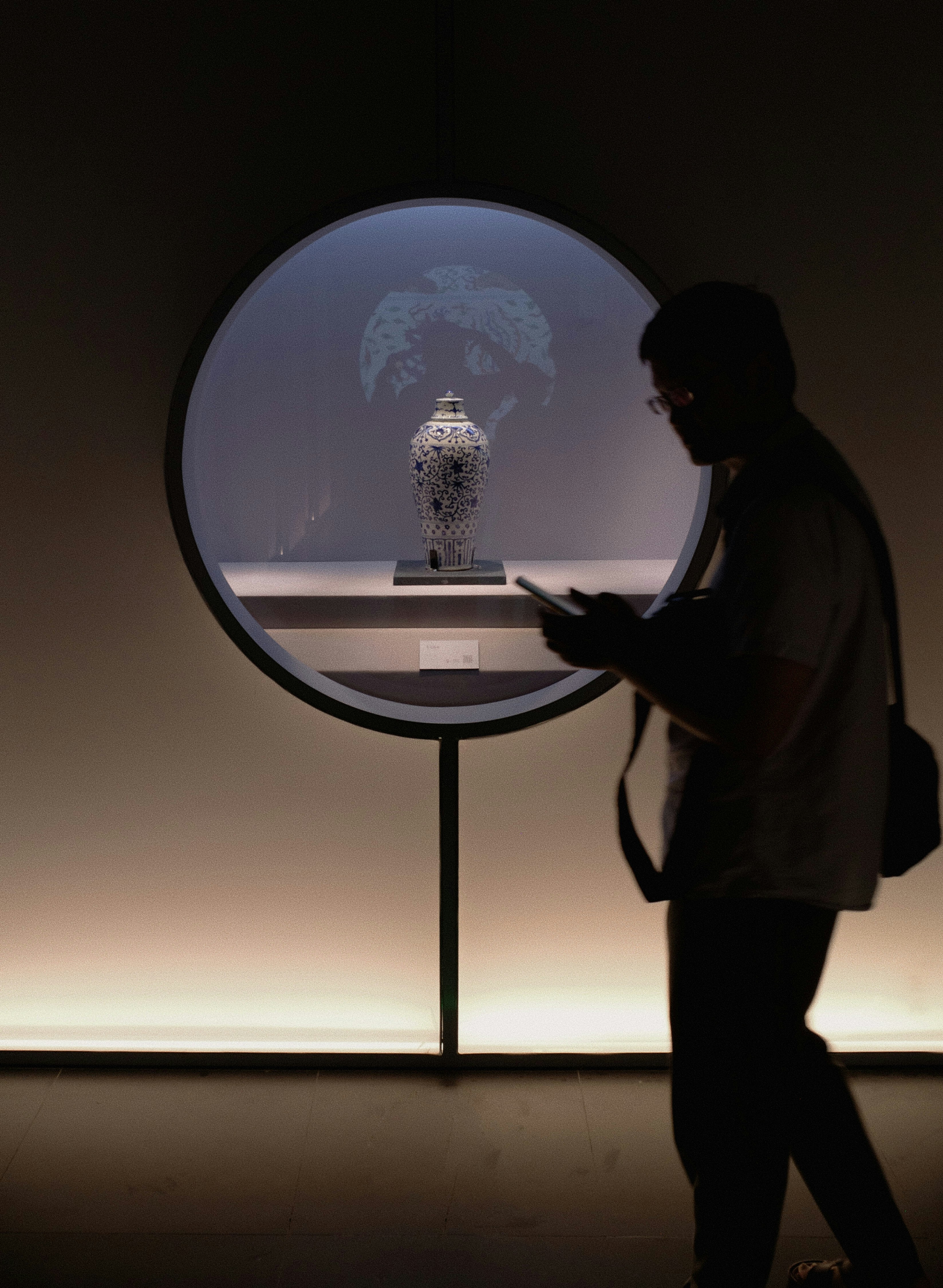 Silhouette of a person passing by an intricately designed vase illuminated within a circular display. The subtle lighting enhances the vase's details.