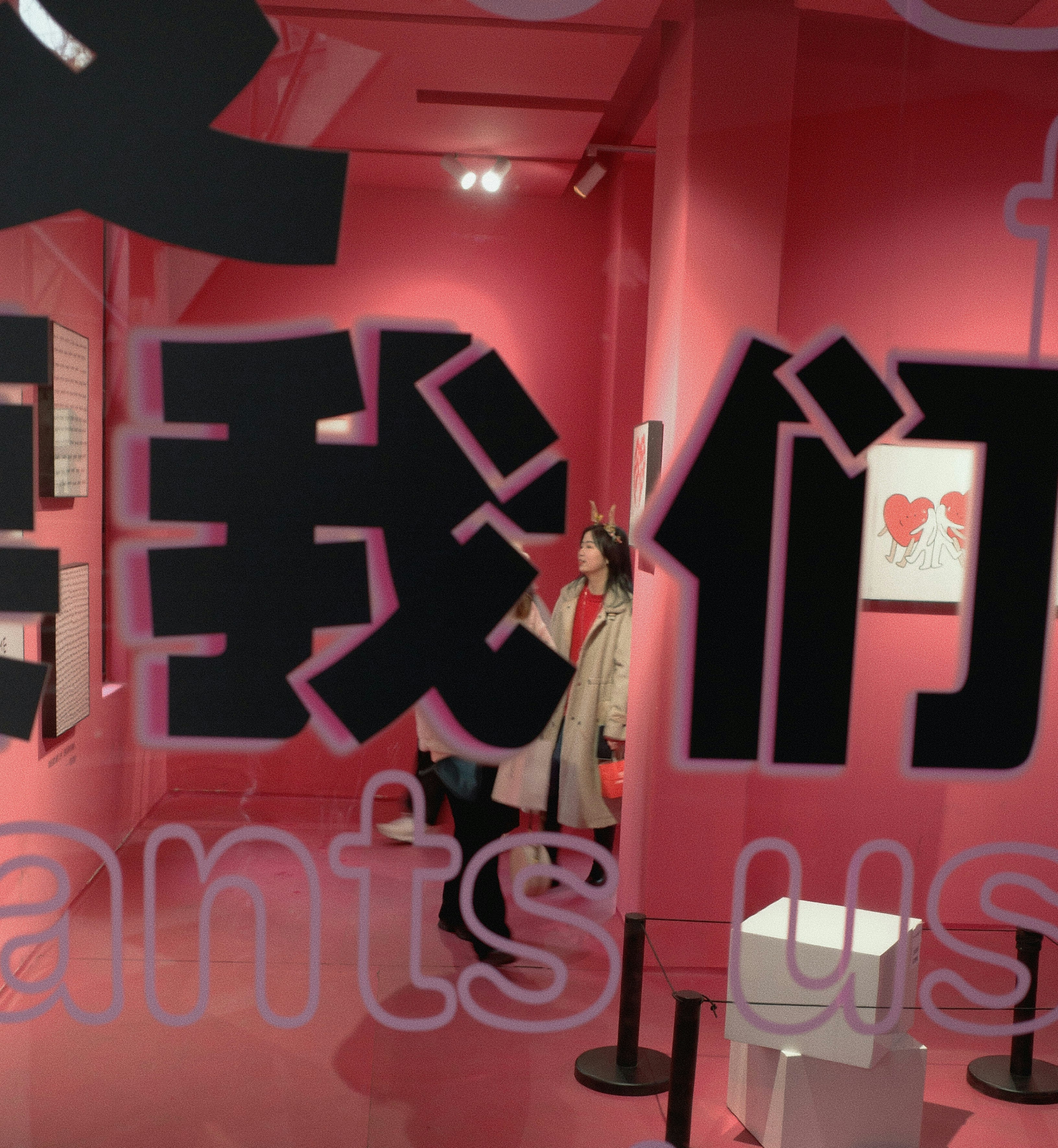A woman in a beige coat observes artwork in a vibrant pink gallery, framed by bold text on glass. The scene captures a moment of engagement with art.
