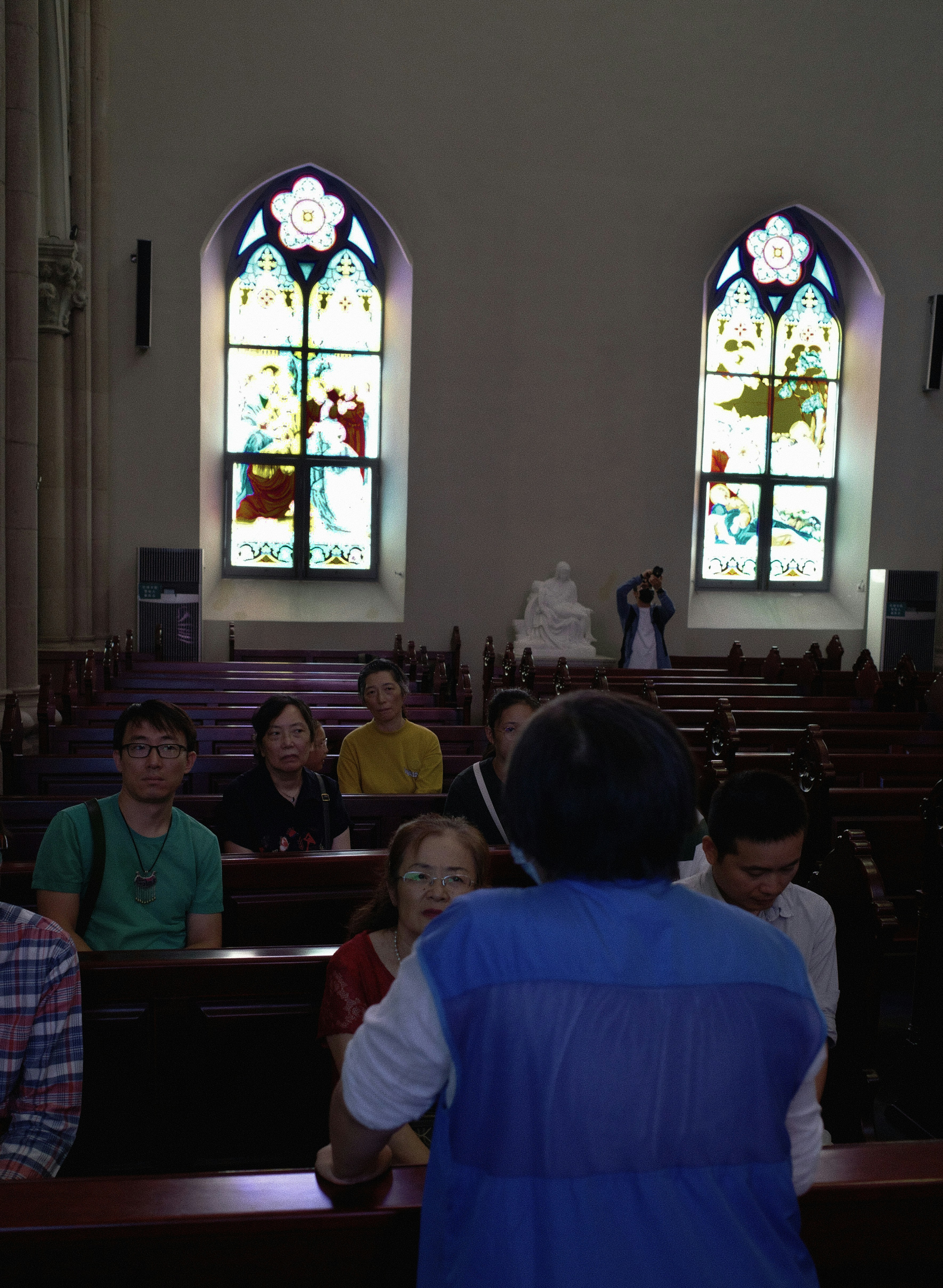 Visitors engaged in contemplation within a church, illuminated by colorful stained glass windows. A figure in blue leads the gathering.